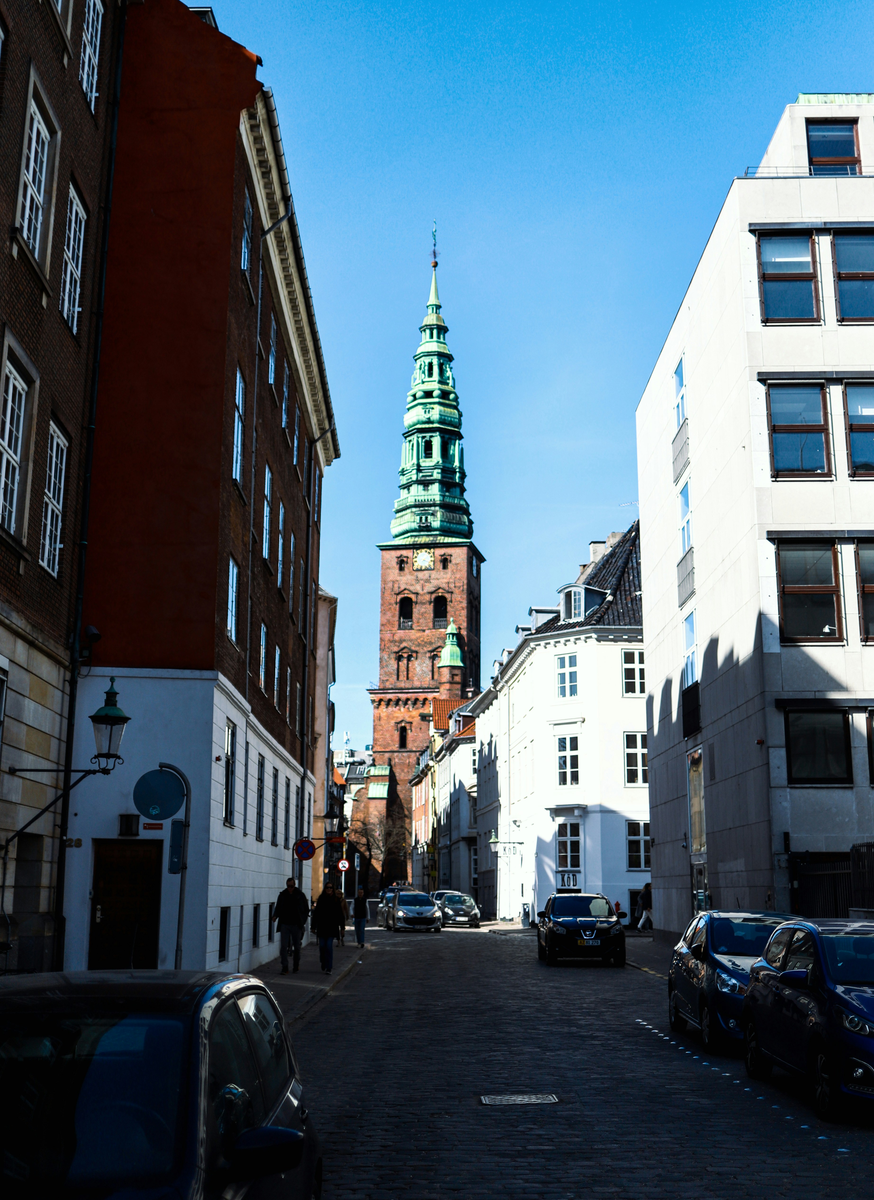 Historic church tower rises above narrow cobblestone street, framed by modern architecture and clear blue sky.