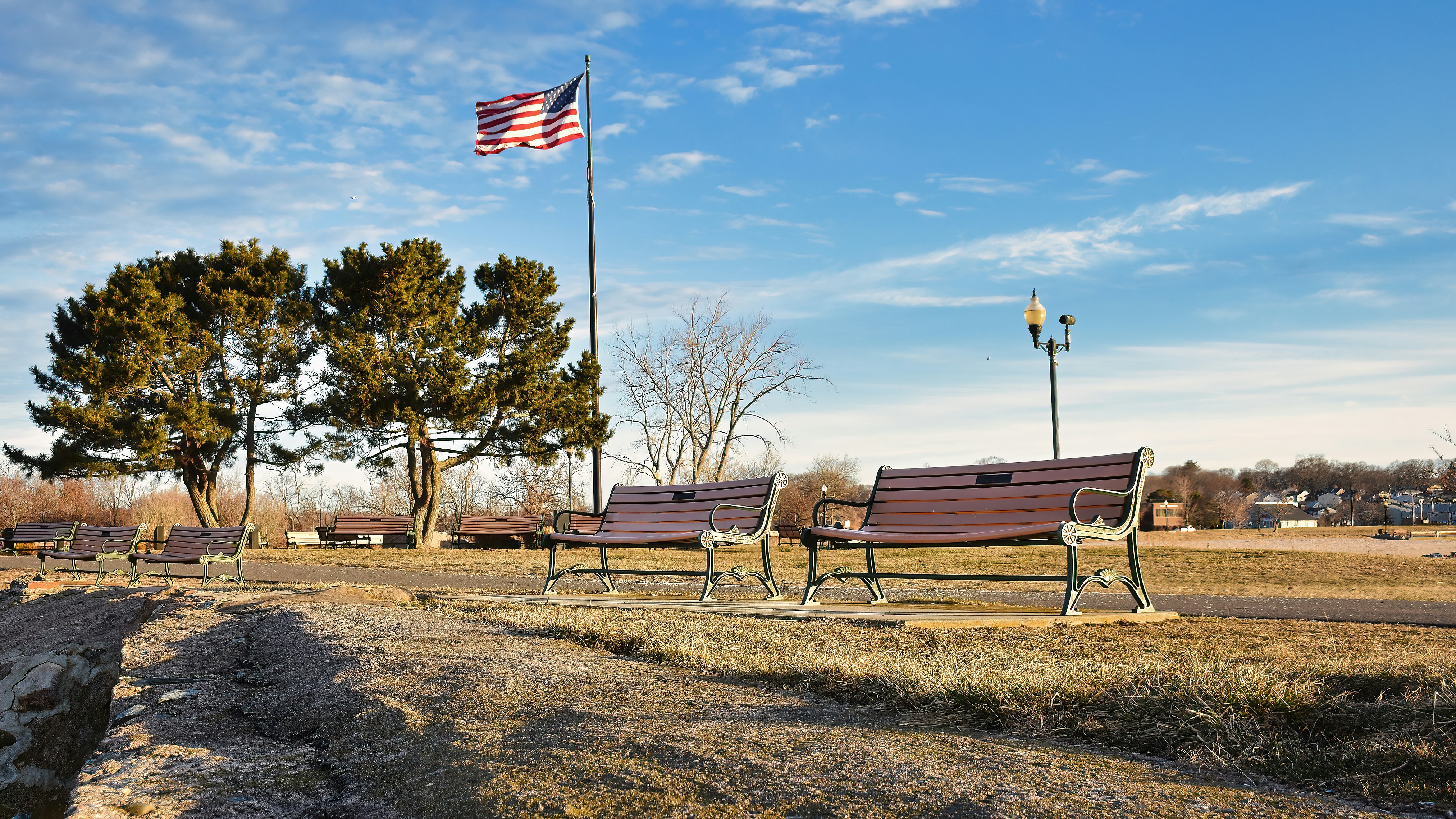 Park benches line a scenic waterfront, framed by trees and an American flag waving in the breeze.