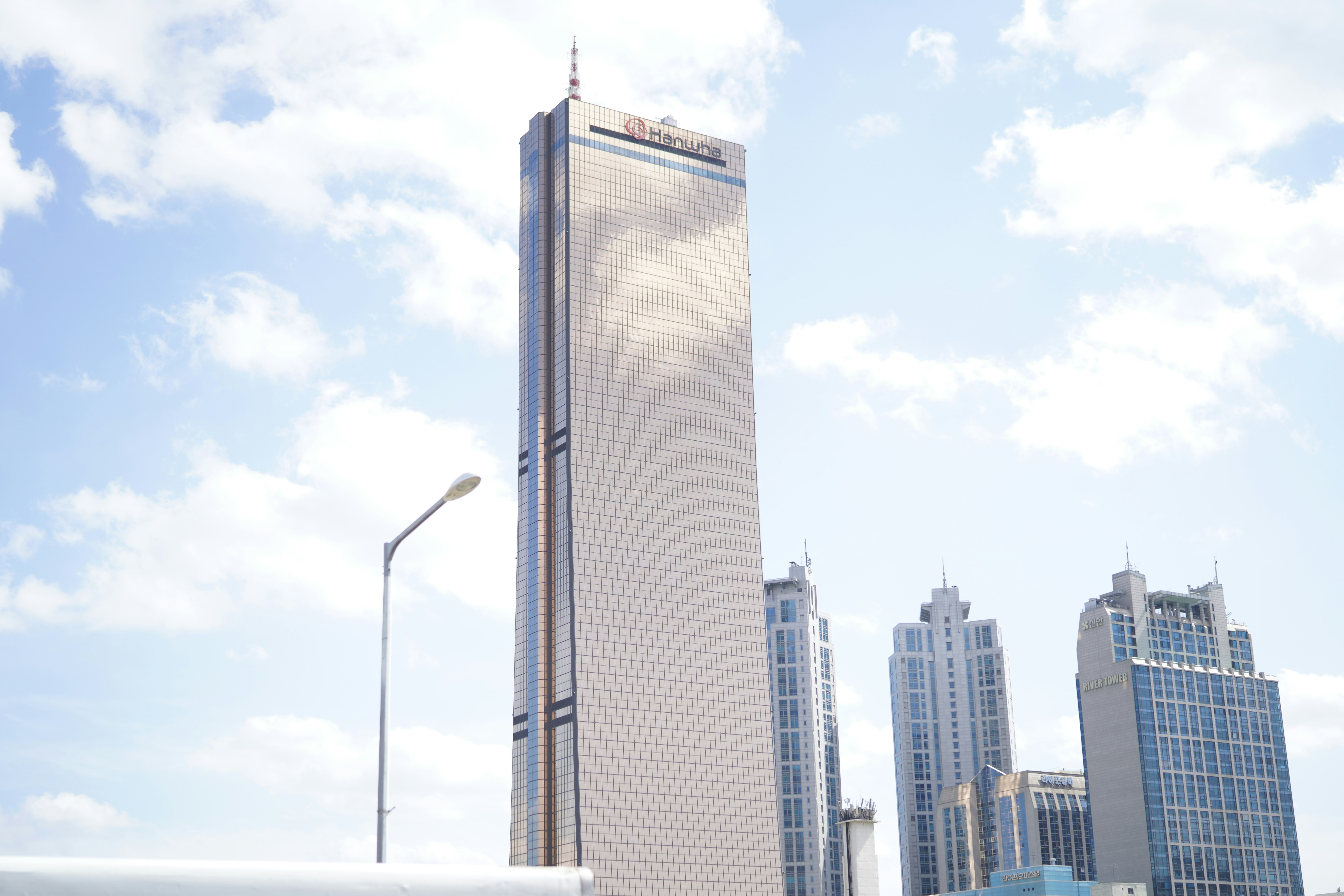 Tall skyscraper reflecting clouds against a bright blue sky, surrounded by modern buildings. The scene conveys urban grandeur.