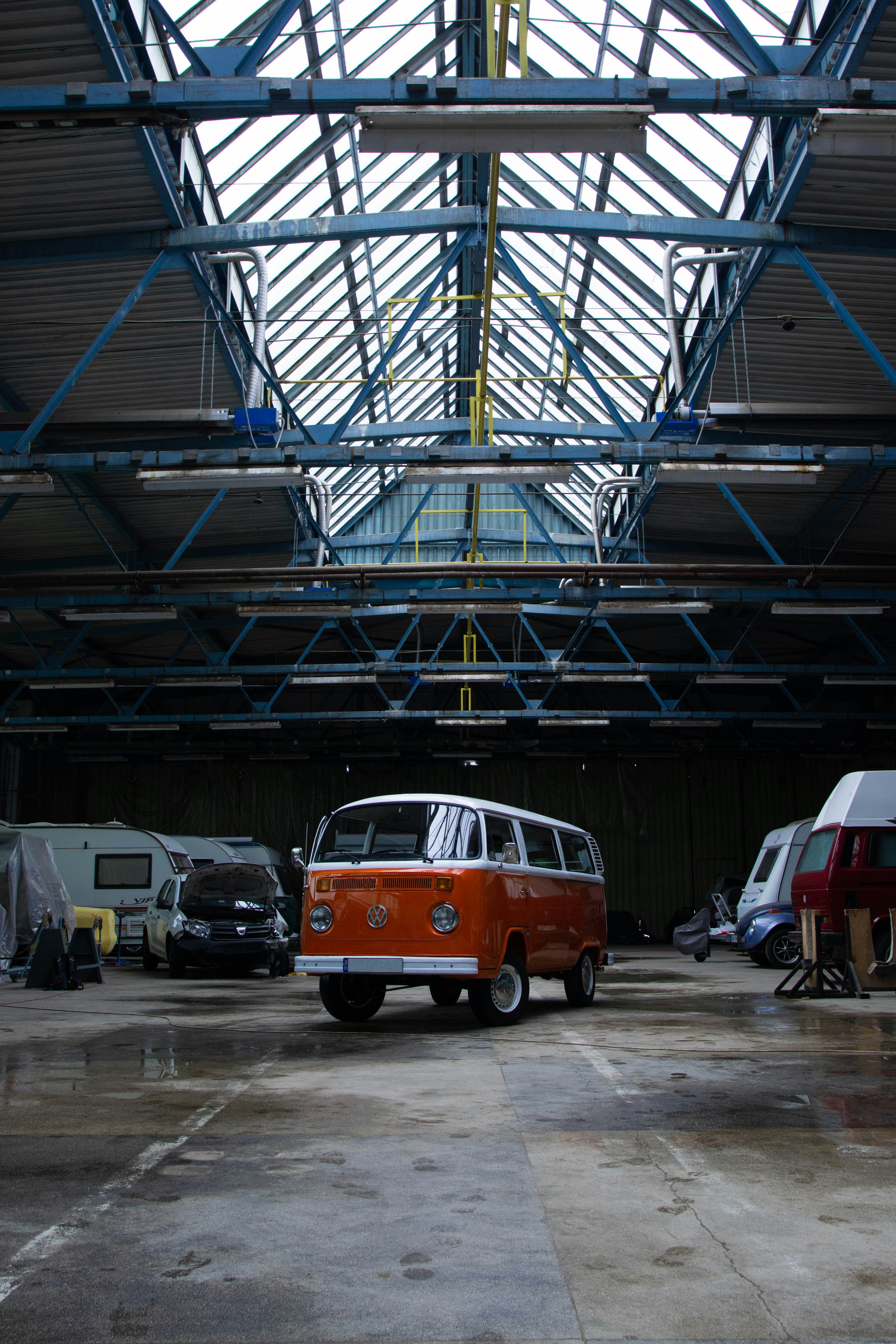 Vintage orange Volkswagen van parked in a spacious industrial garage under a skylight, surrounded by various vehicles.