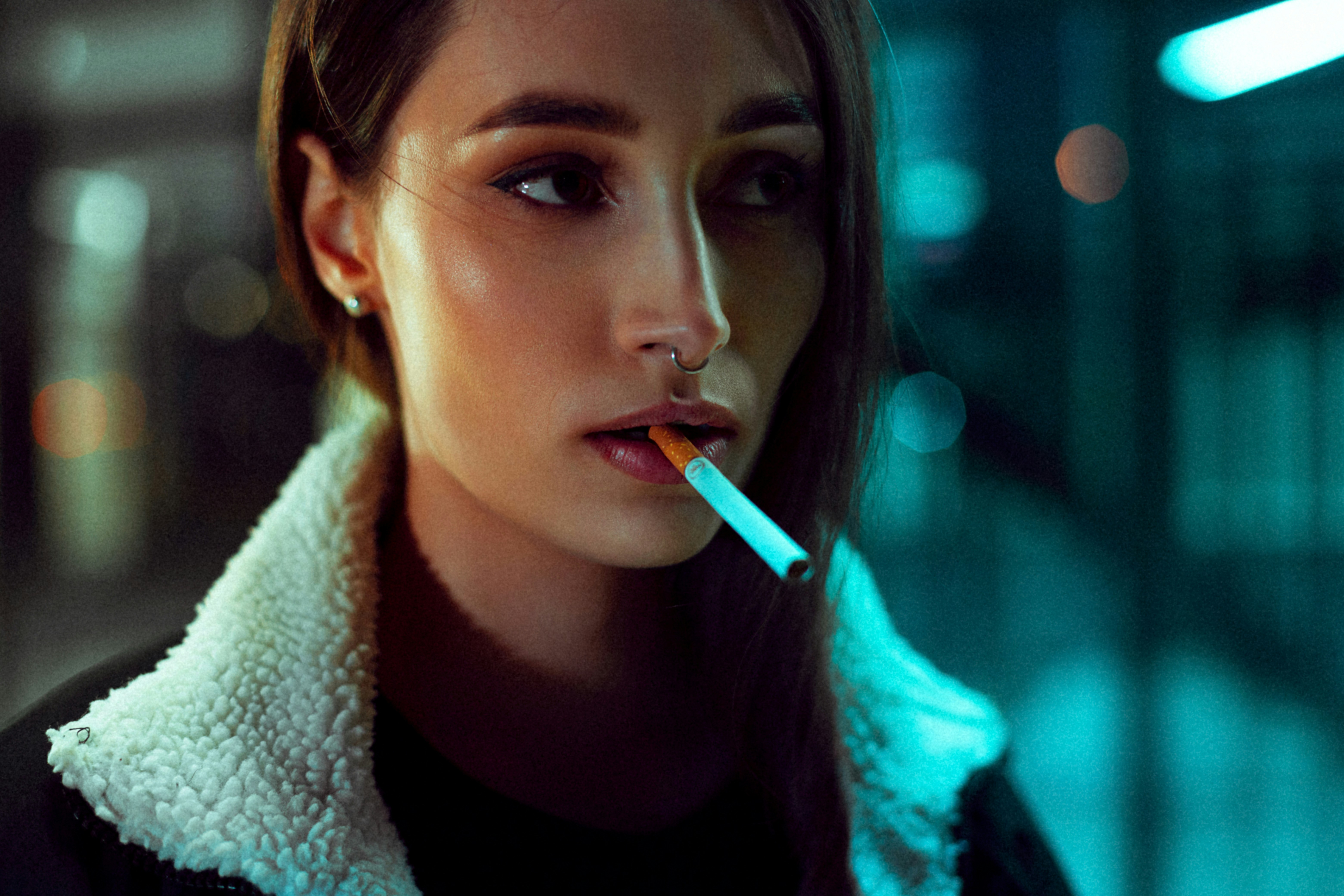 A young woman with a cigarette in her mouth gazes pensively, surrounded by a softly lit urban backdrop. The contrast of her expression and the ambient light creates a moody atmosphere.