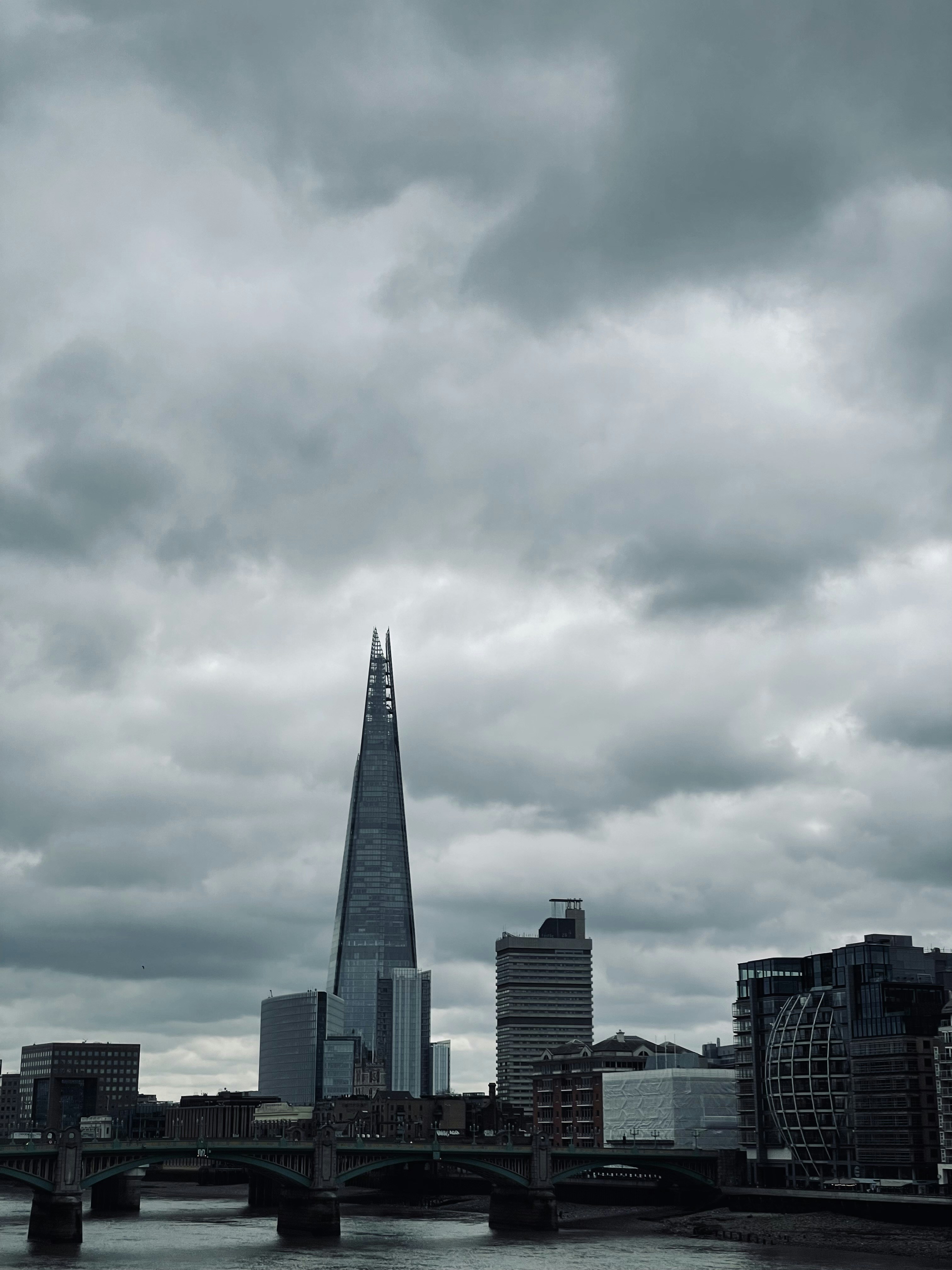 The Shard skyscraper rises dramatically against a backdrop of moody clouds, framed by the River Thames and urban architecture.