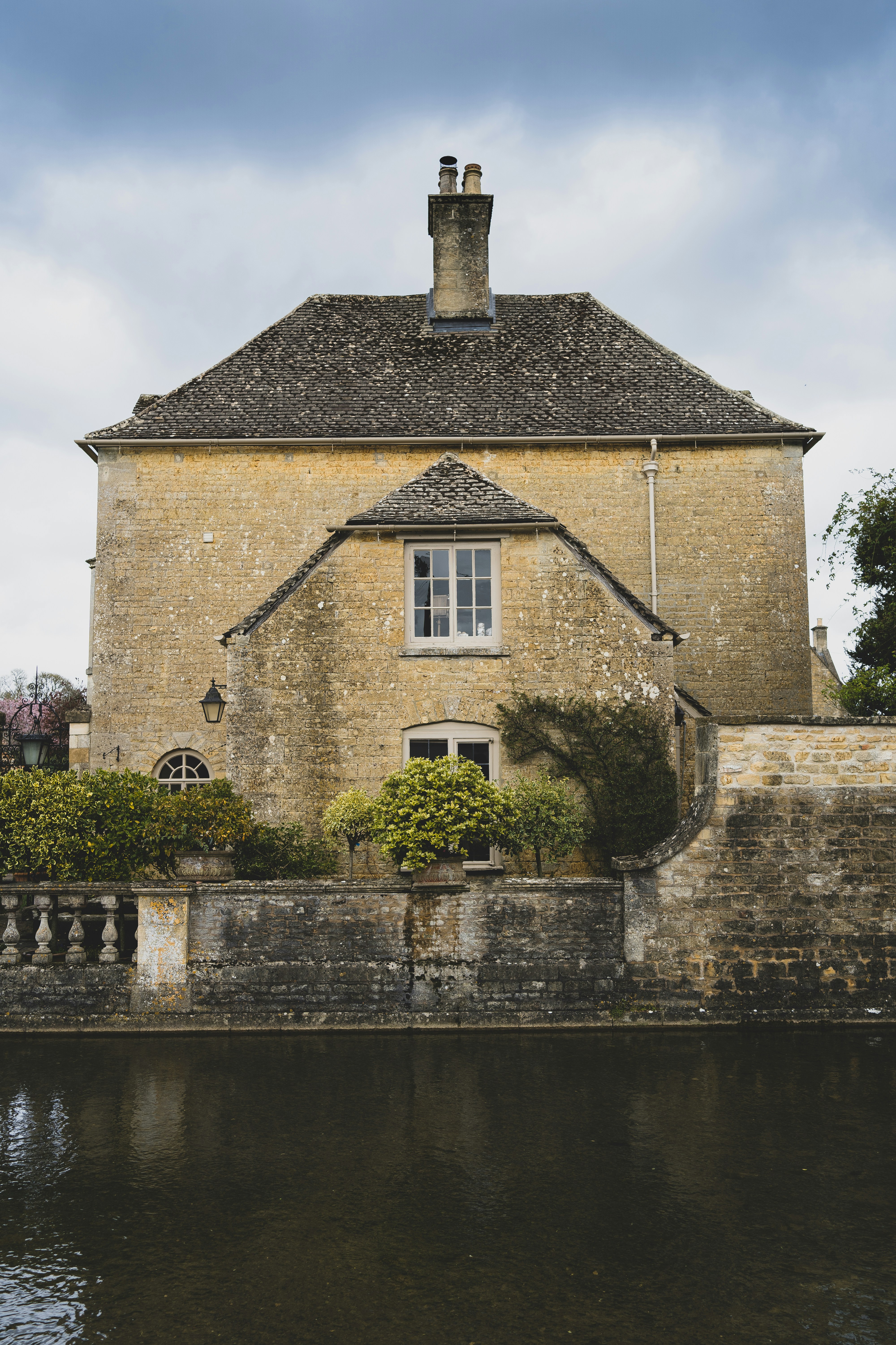 Charming stone cottage with a slate roof, surrounded by lush greenery and reflected in the calm water below. A serene atmosphere invites exploration.