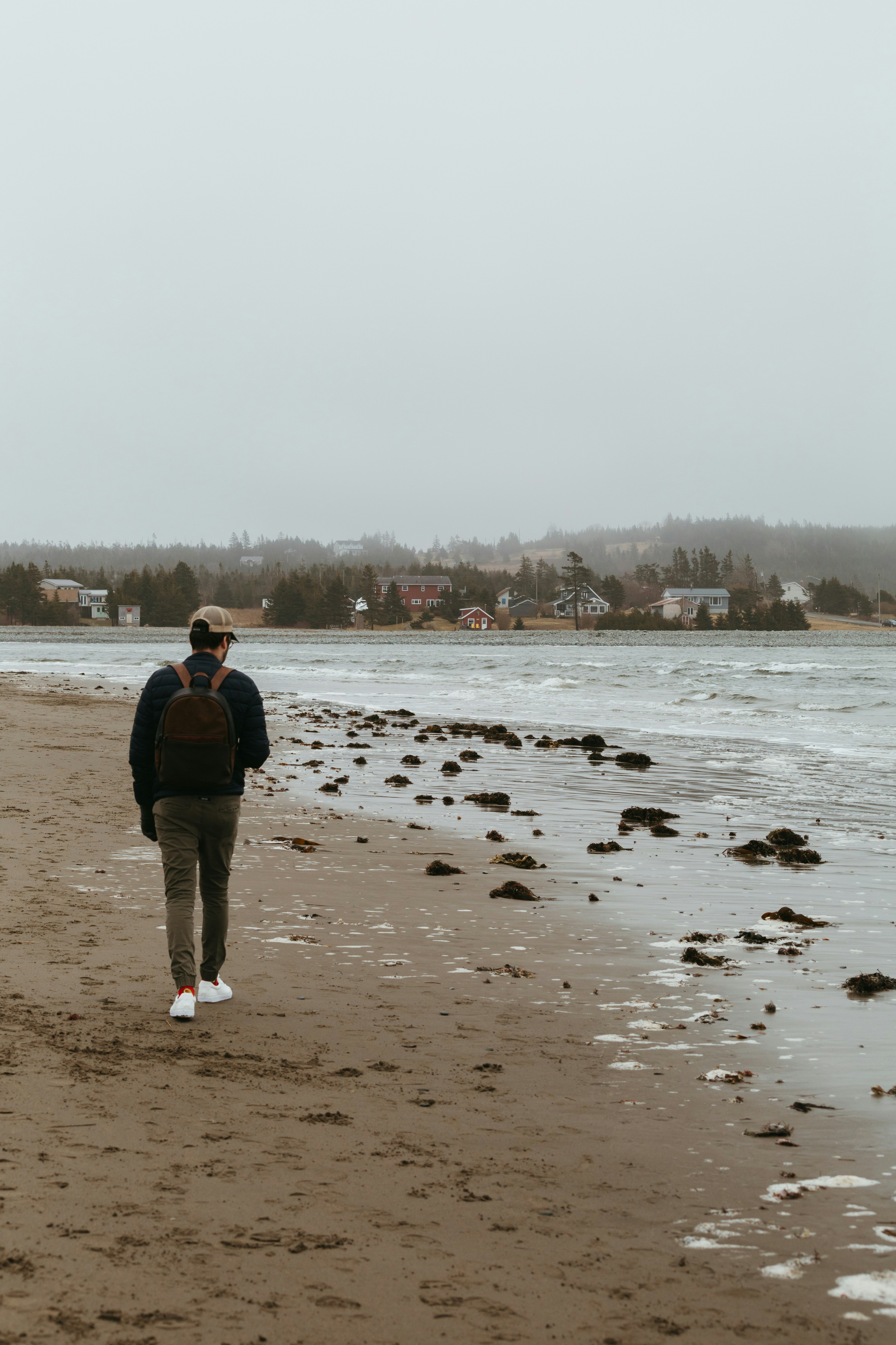 A solitary figure walks along a sandy beach, with gentle waves lapping at the shore and foggy coastal homes in the background.