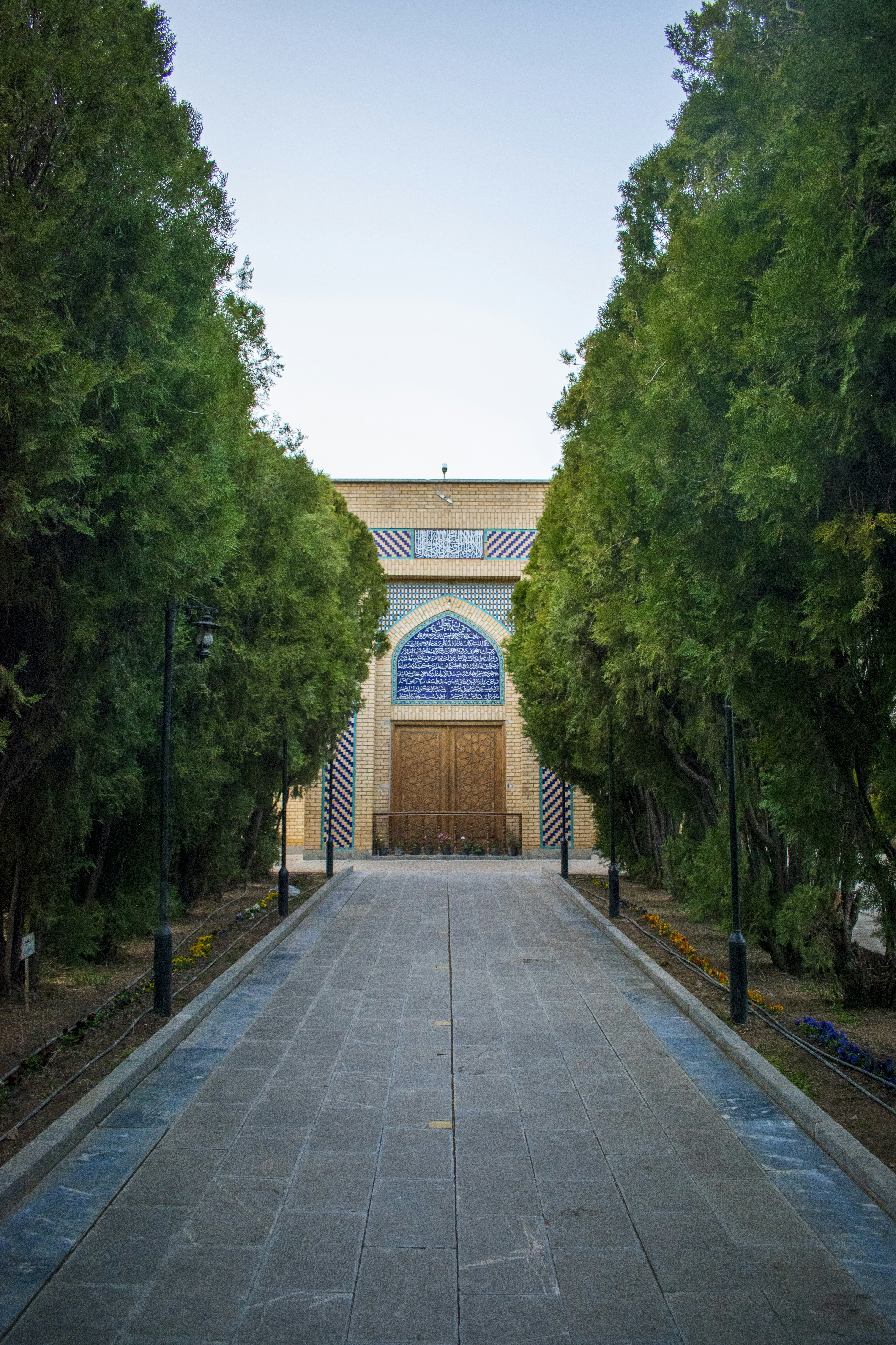 A tranquil pathway bordered by lush trees leading to an ornate entrance with intricate tile work. The scene invites contemplation and exploration.