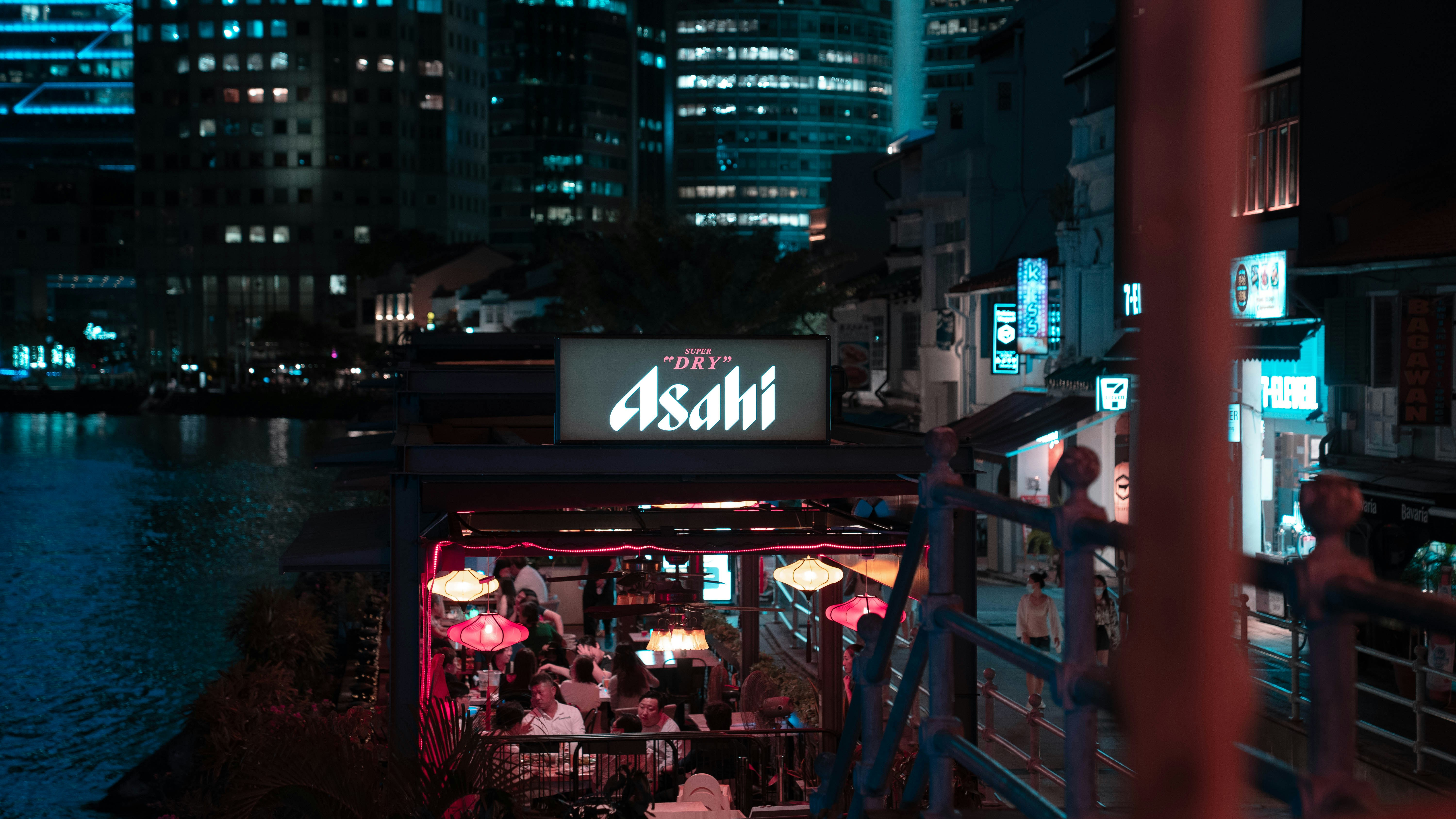 A vibrant riverside restaurant illuminated by colorful neon lights, with patrons enjoying their evening. The Asahi sign prominently stands out against the urban backdrop.