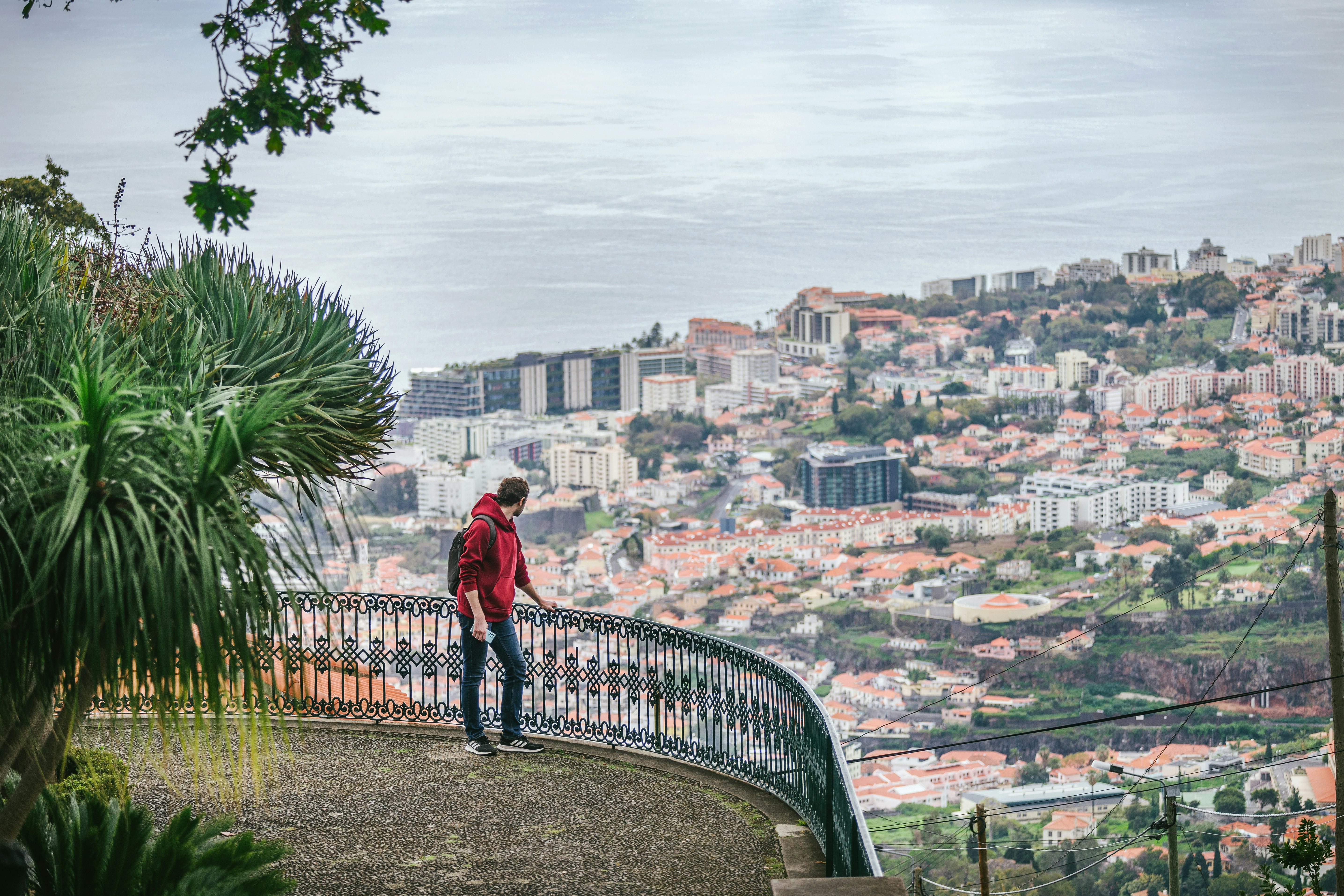 A lone figure in a red hoodie stands on a curved railing, overlooking a sprawling cityscape dotted with terracotta roofs and greenery.