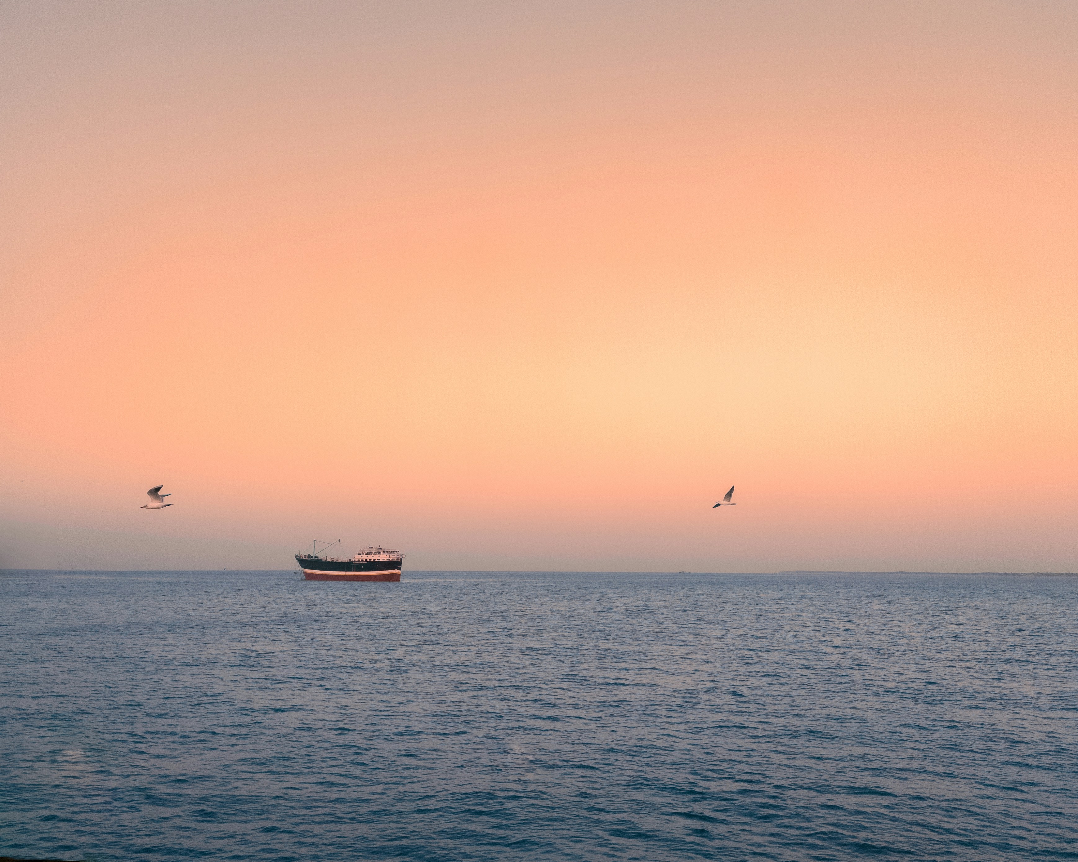 A solitary boat drifts on calm ocean waters under a peach-hued sunrise sky.