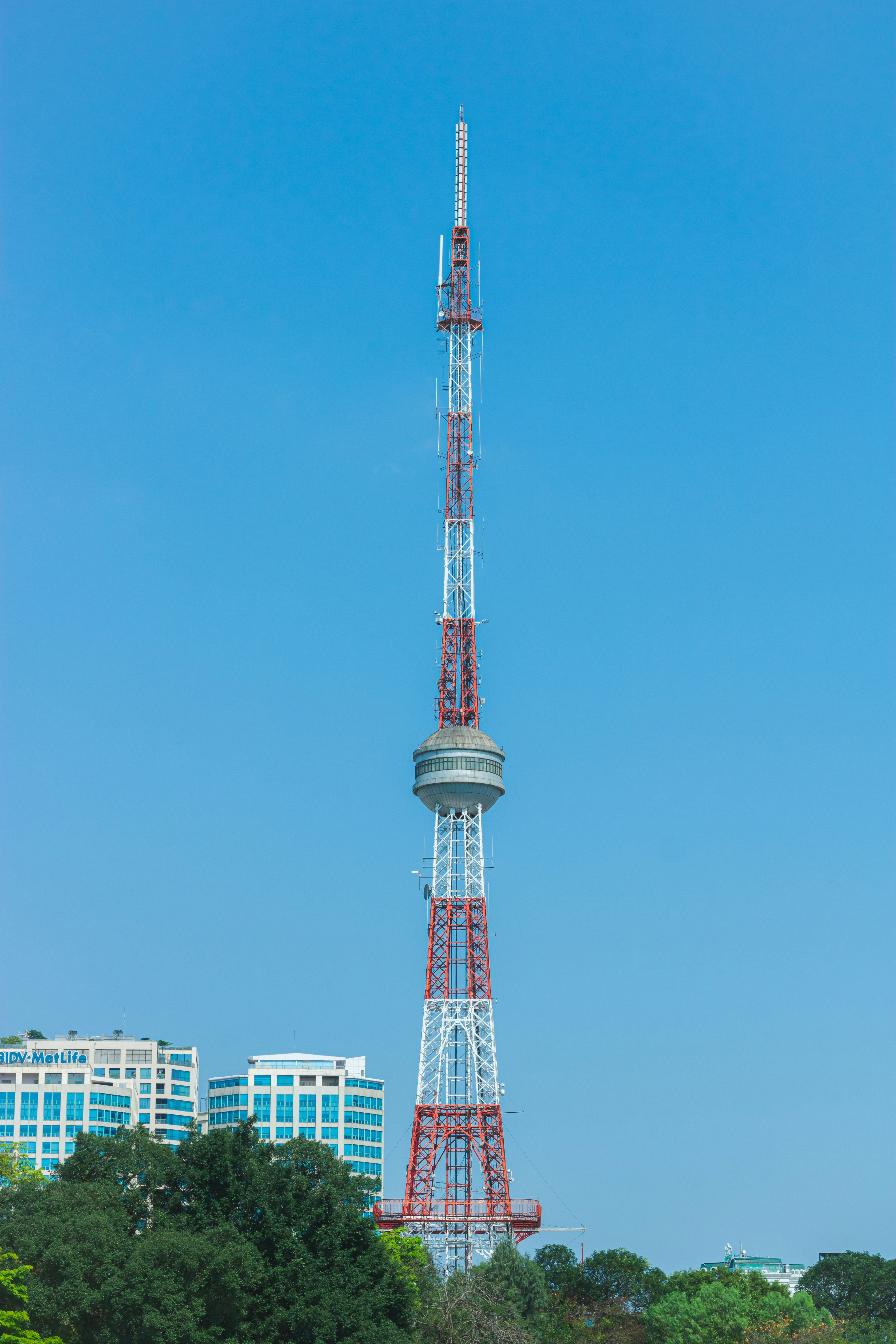 Telecommunication tower rising against a clear blue sky, surrounded by modern buildings and lush greenery.