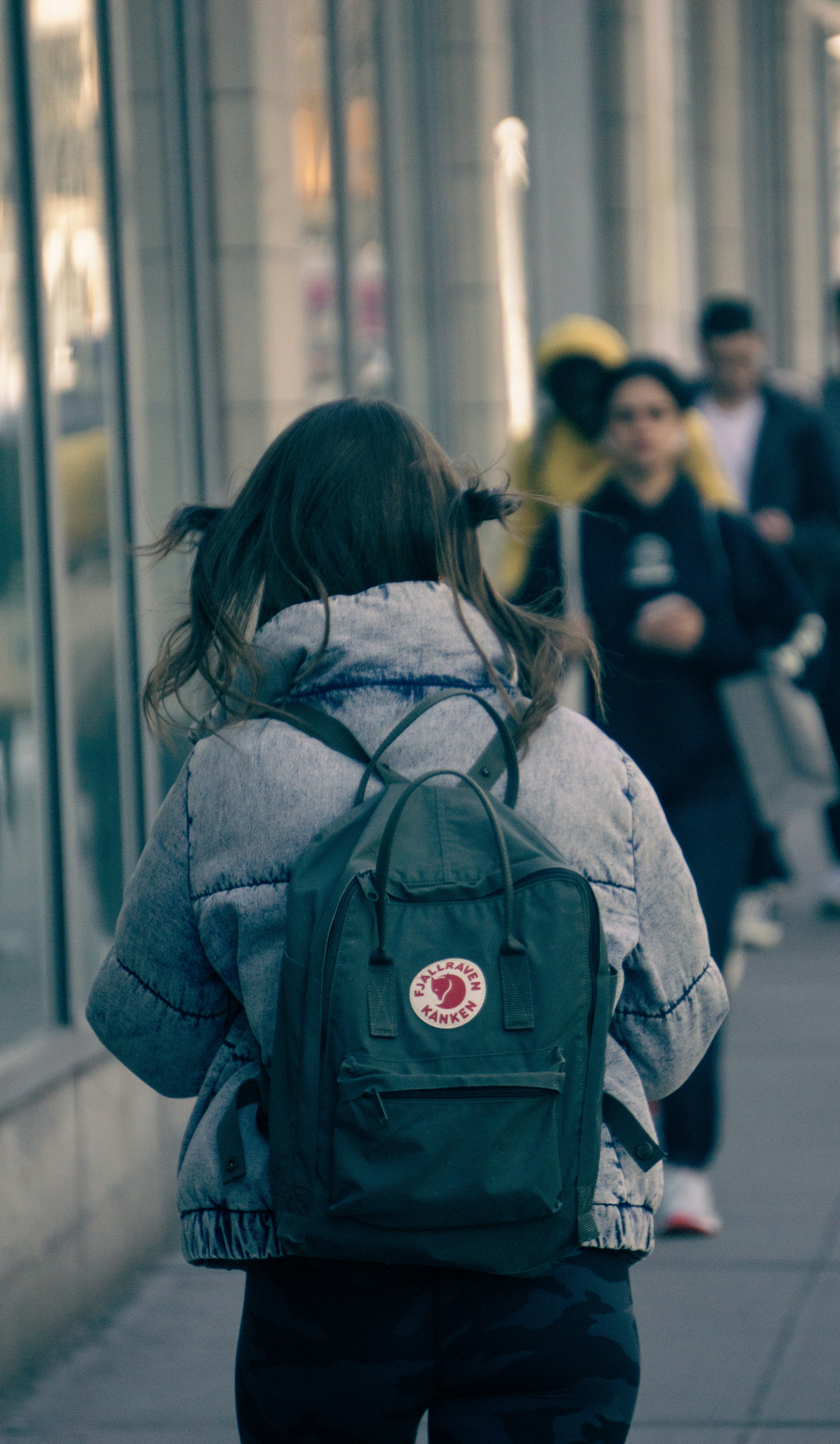 A young woman with a backpack walks through a bustling city street, her hair caught in the breeze, as blurred figures pass by in the background.