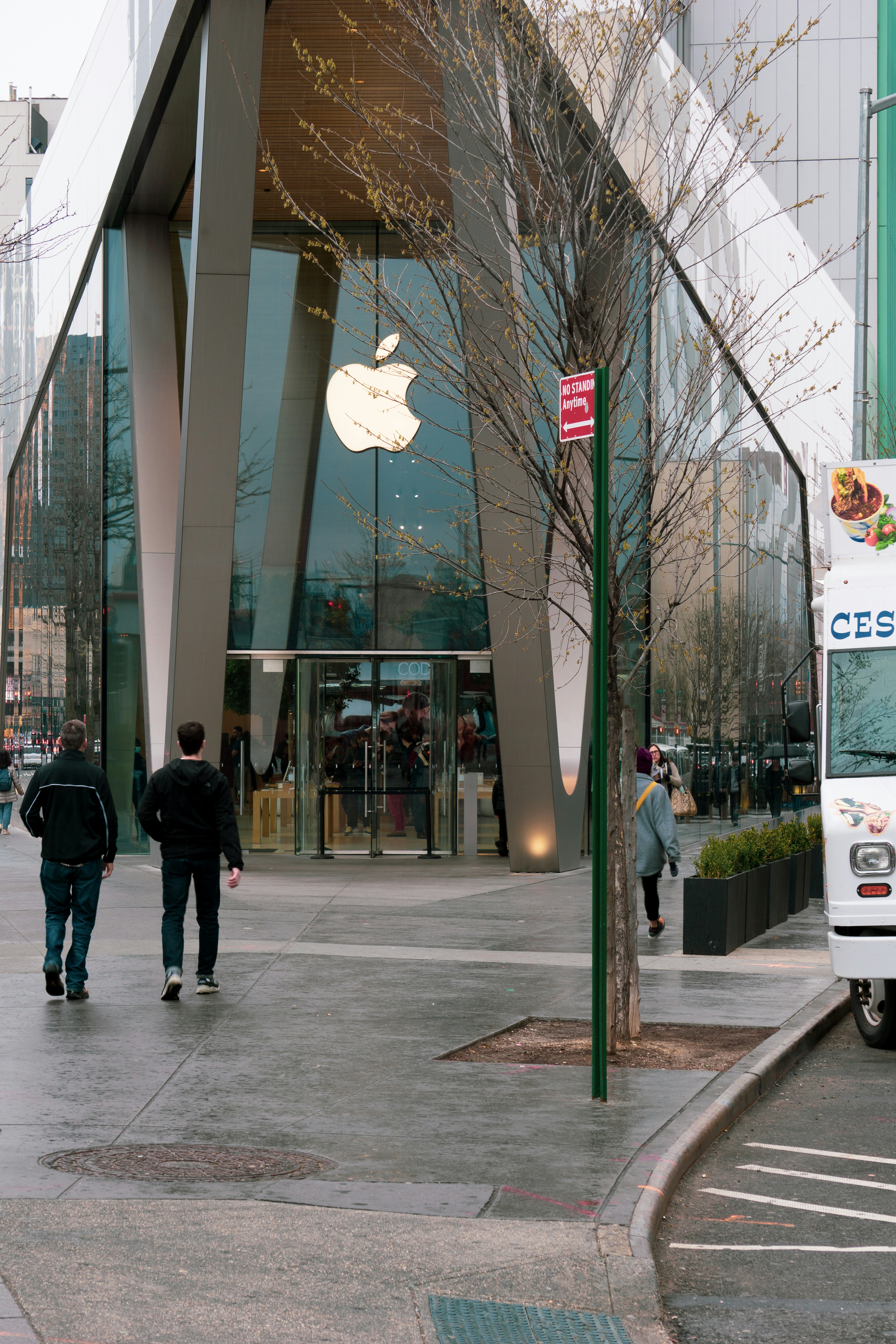 Modern Apple Store entrance framed by sleek architecture, with pedestrians walking by and a food truck in view.
