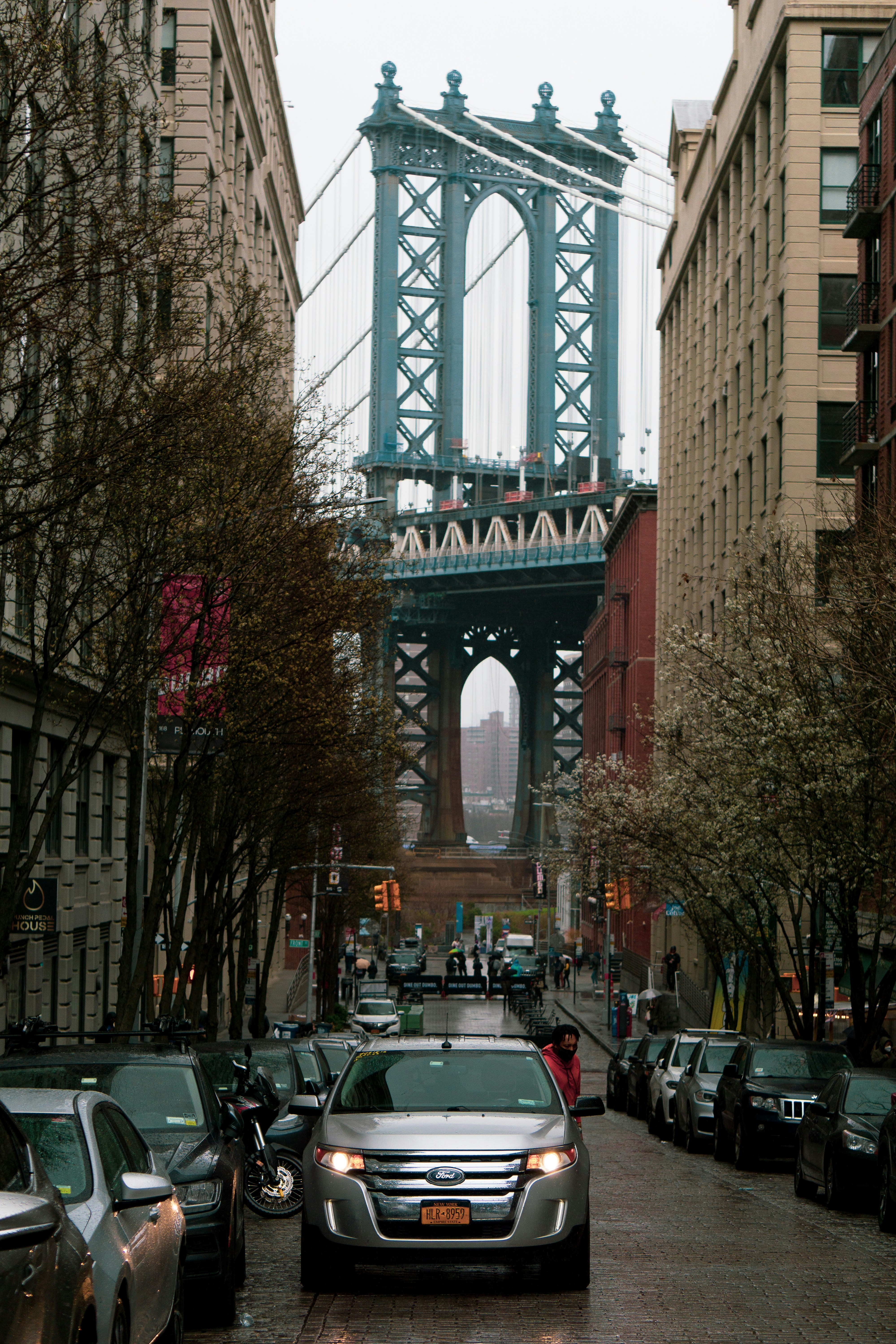 A busy street scene framed by buildings, leading to the iconic Manhattan Bridge, with a car in the foreground and pedestrians navigating the urban landscape.