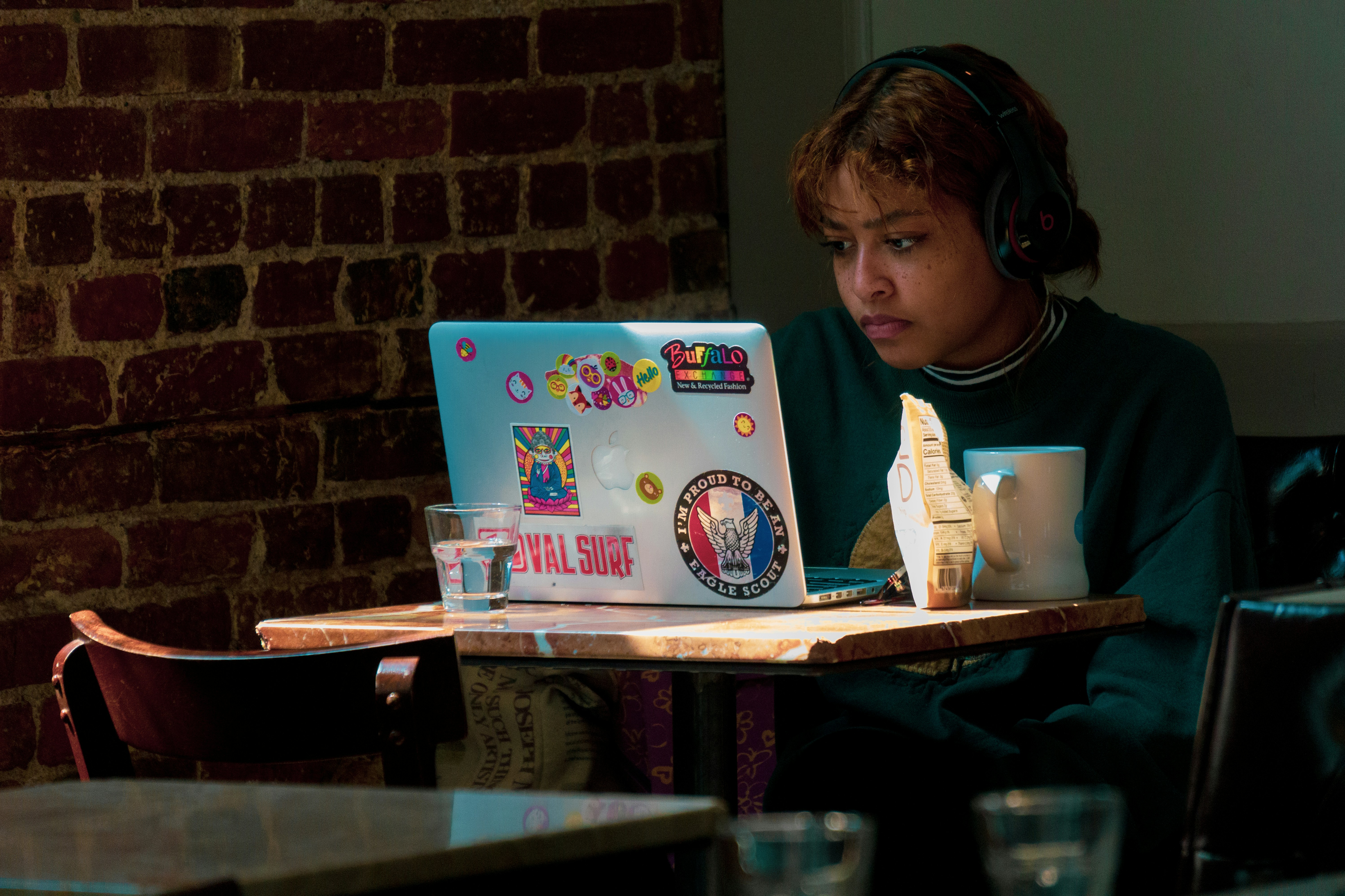 Individual immersed in work at a café, surrounded by a brick wall and soft lighting, with a laptop covered in colorful stickers. A cup and snacks are nearby.