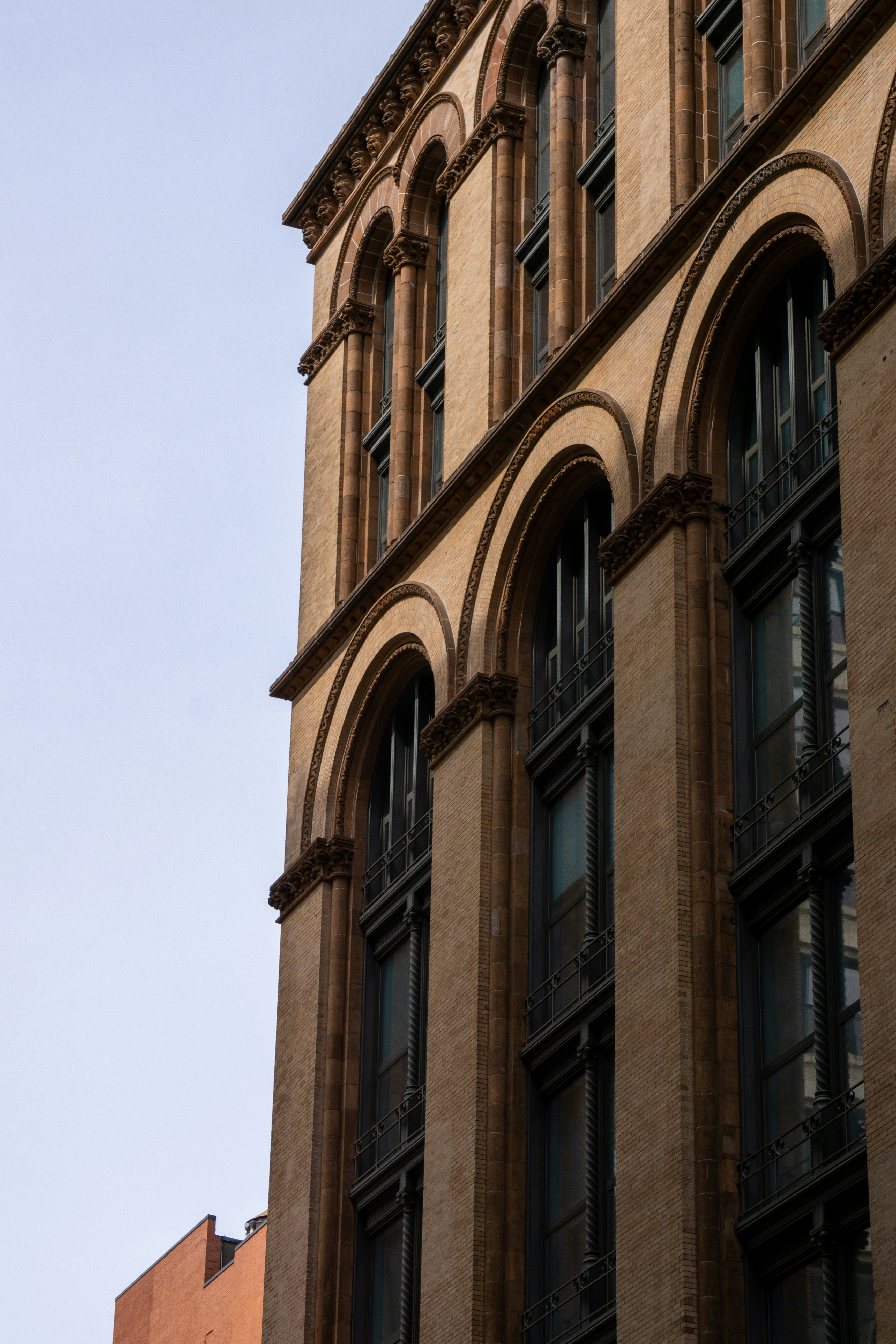 Detailed view of a historic building's arched windows and textured brickwork against a soft sky. The composition highlights the architectural intricacies and urban charm.
