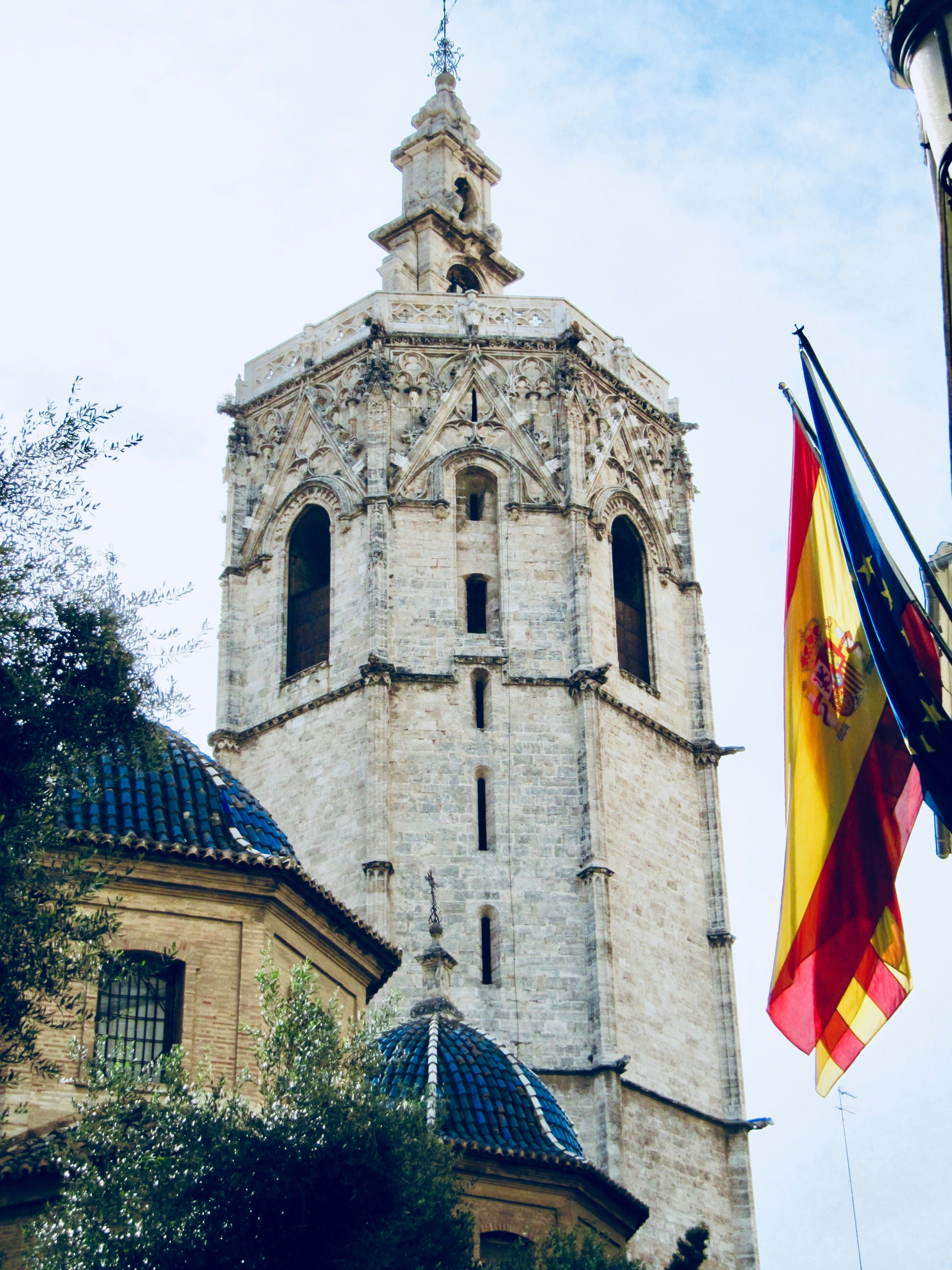 Gothic bell tower rising majestically against a blue sky, flanked by vibrant flags and lush greenery.