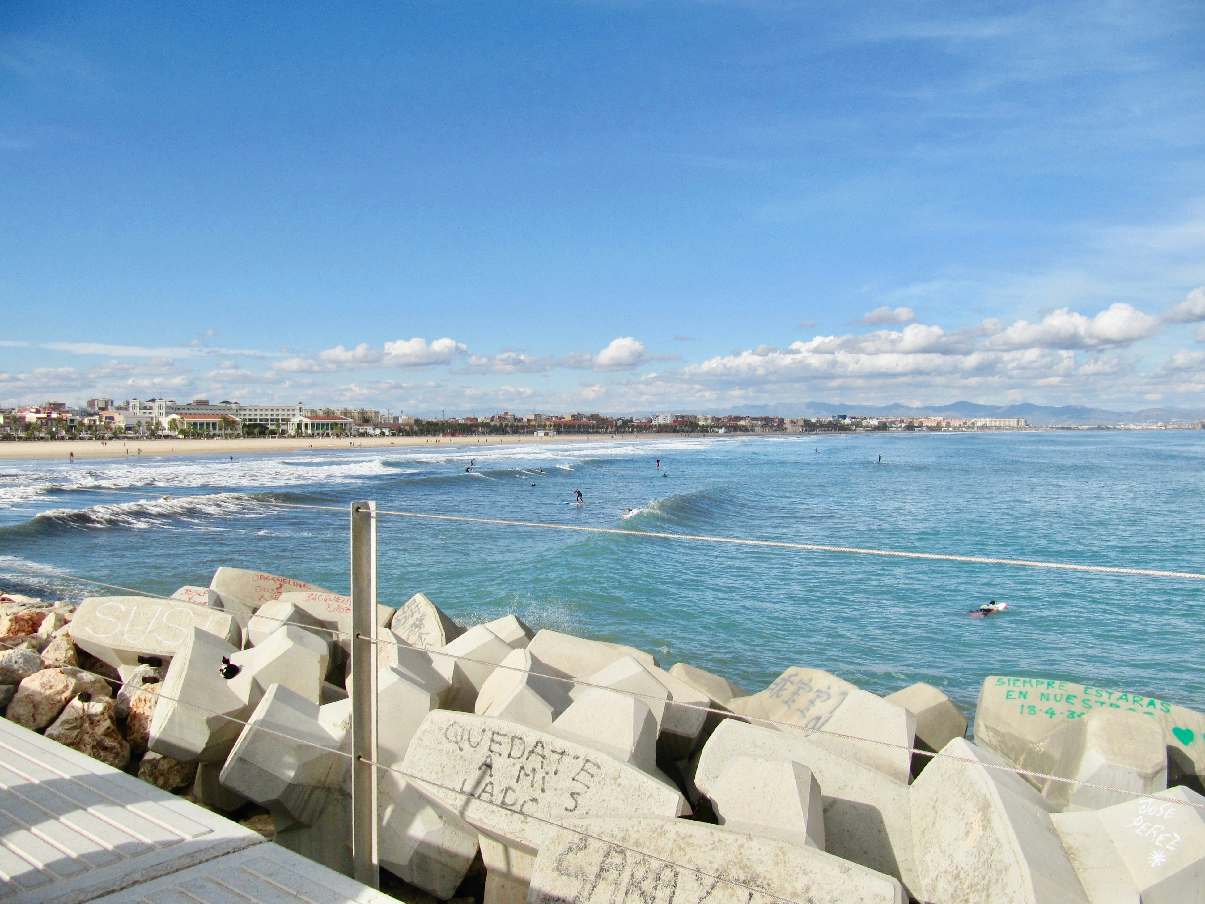 Coastal scene featuring surfers riding gentle waves beside a rocky shoreline with urban buildings in the background. The sky is clear with scattered clouds.