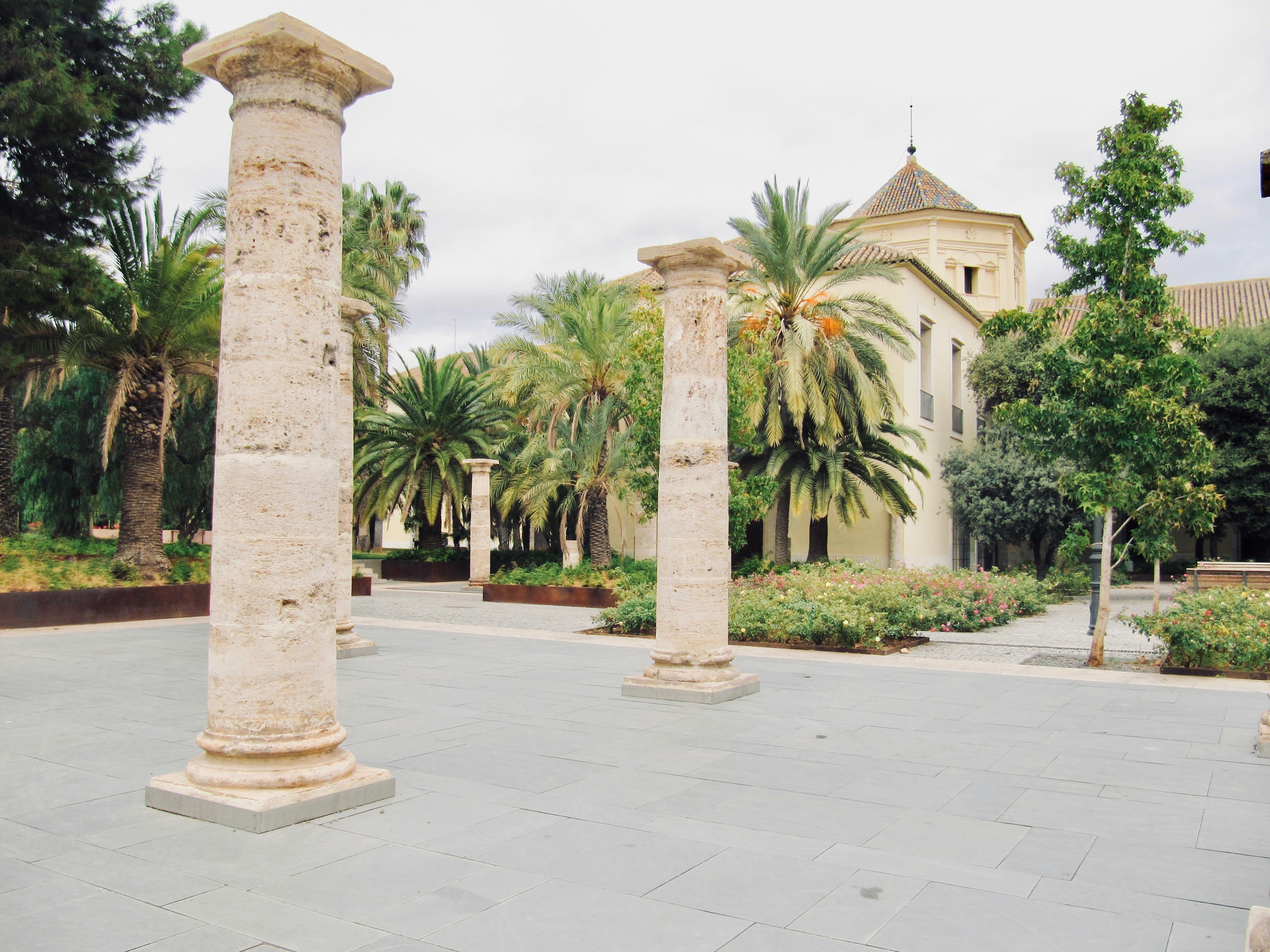 Ancient stone columns stand amidst vibrant palm trees and a historic building, highlighting a blend of nature and architecture.