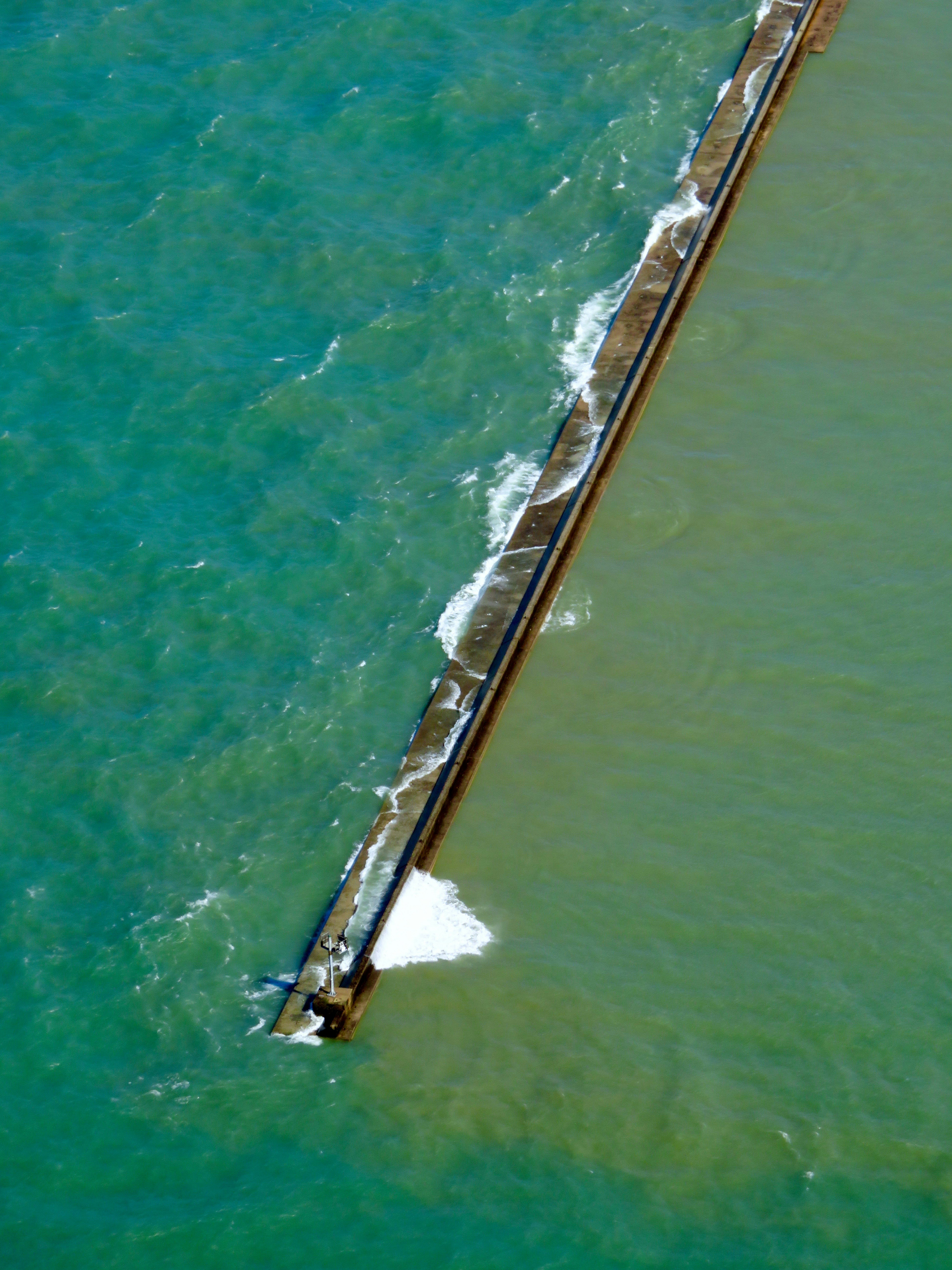 A jetty stretches into the turquoise waters, creating a stark contrast with the deeper hues of the ocean. Waves gently crash against the structure, illustrating the dynamic interaction between land and sea.