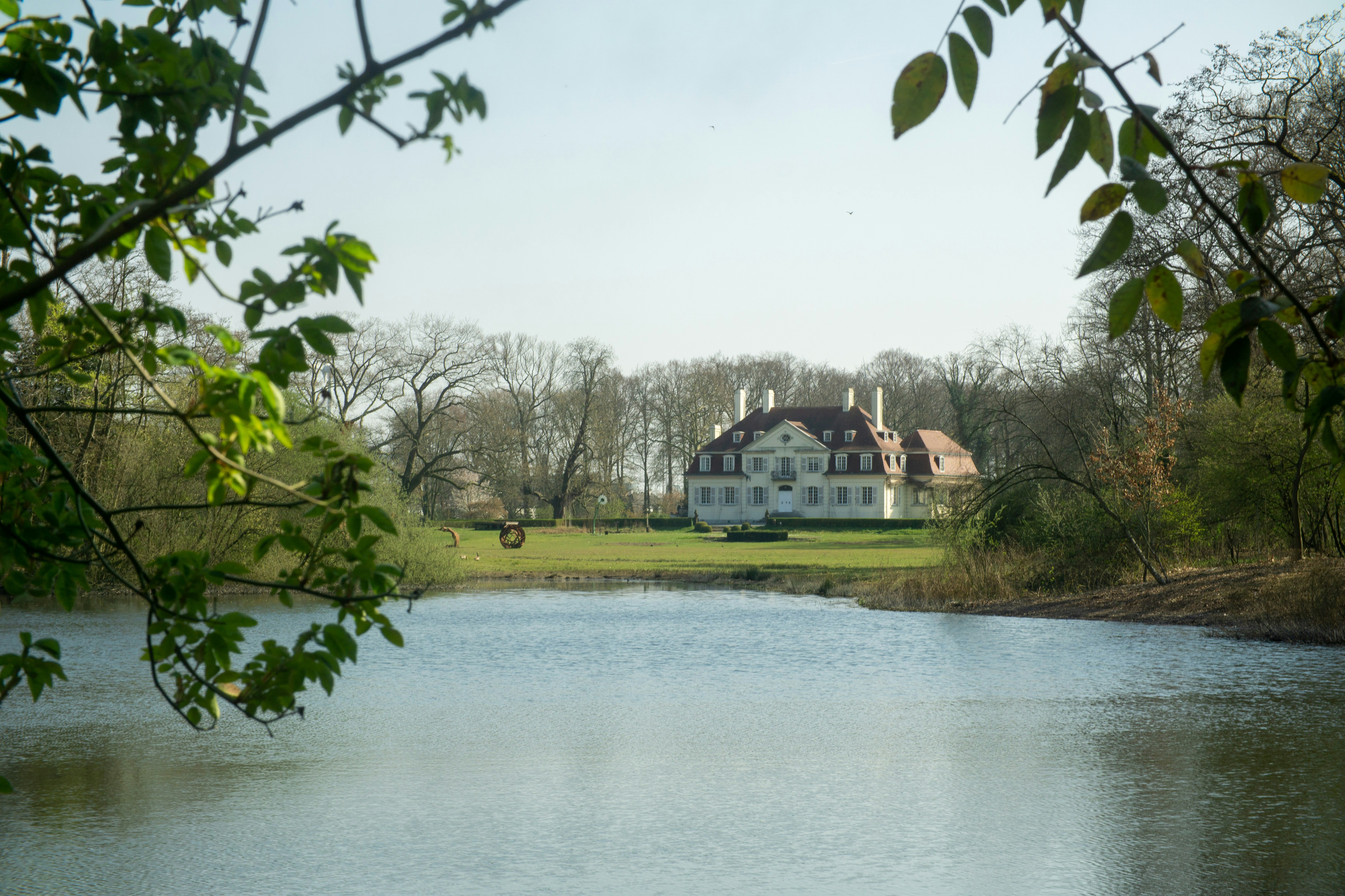 Grand manor framed by leafy branches across a serene lake under a clear sky.