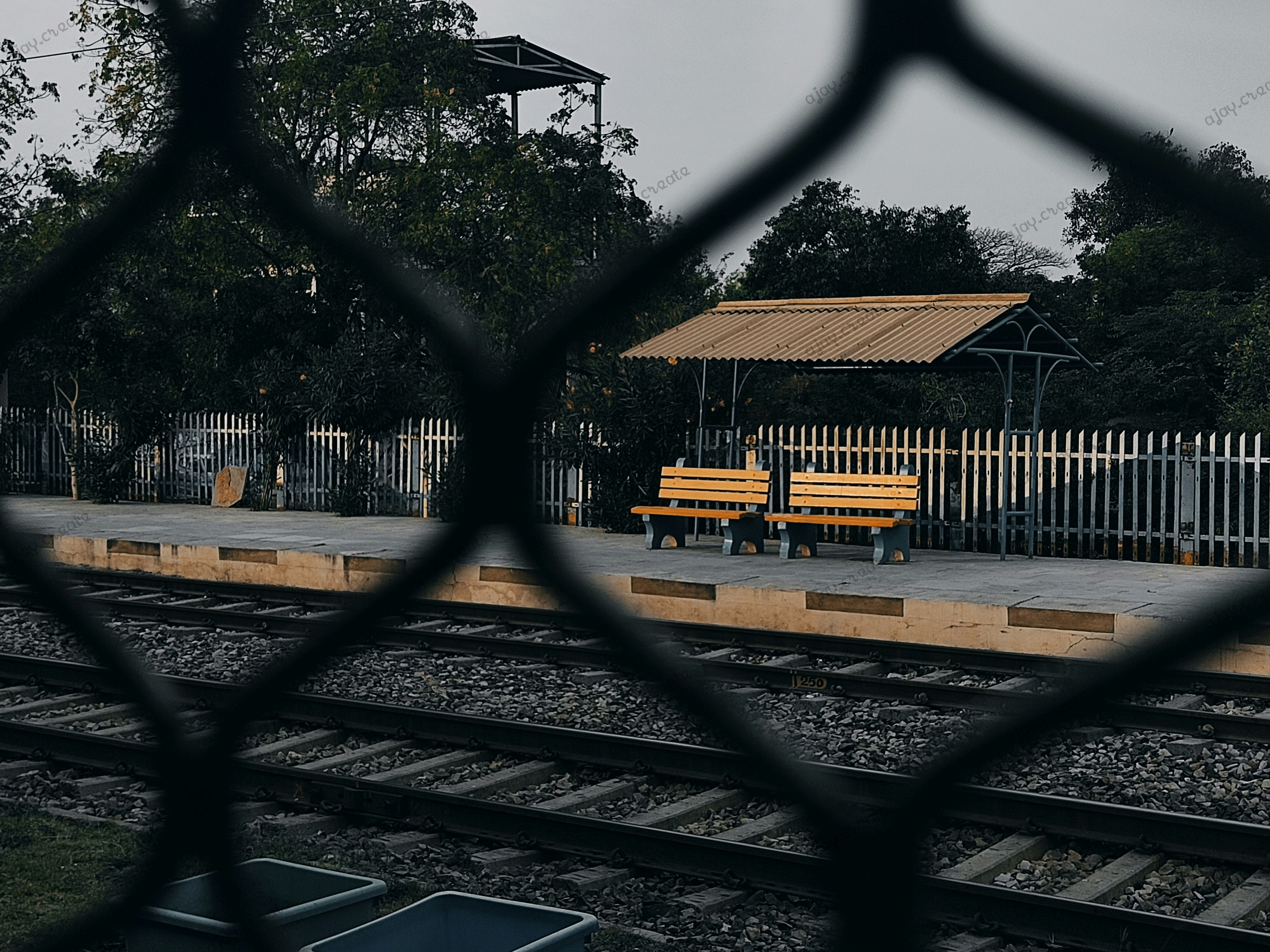 Yellow bench under a rustic shelter at a train station, framed by a chain-link fence. The scene evokes a sense of solitude and anticipation.