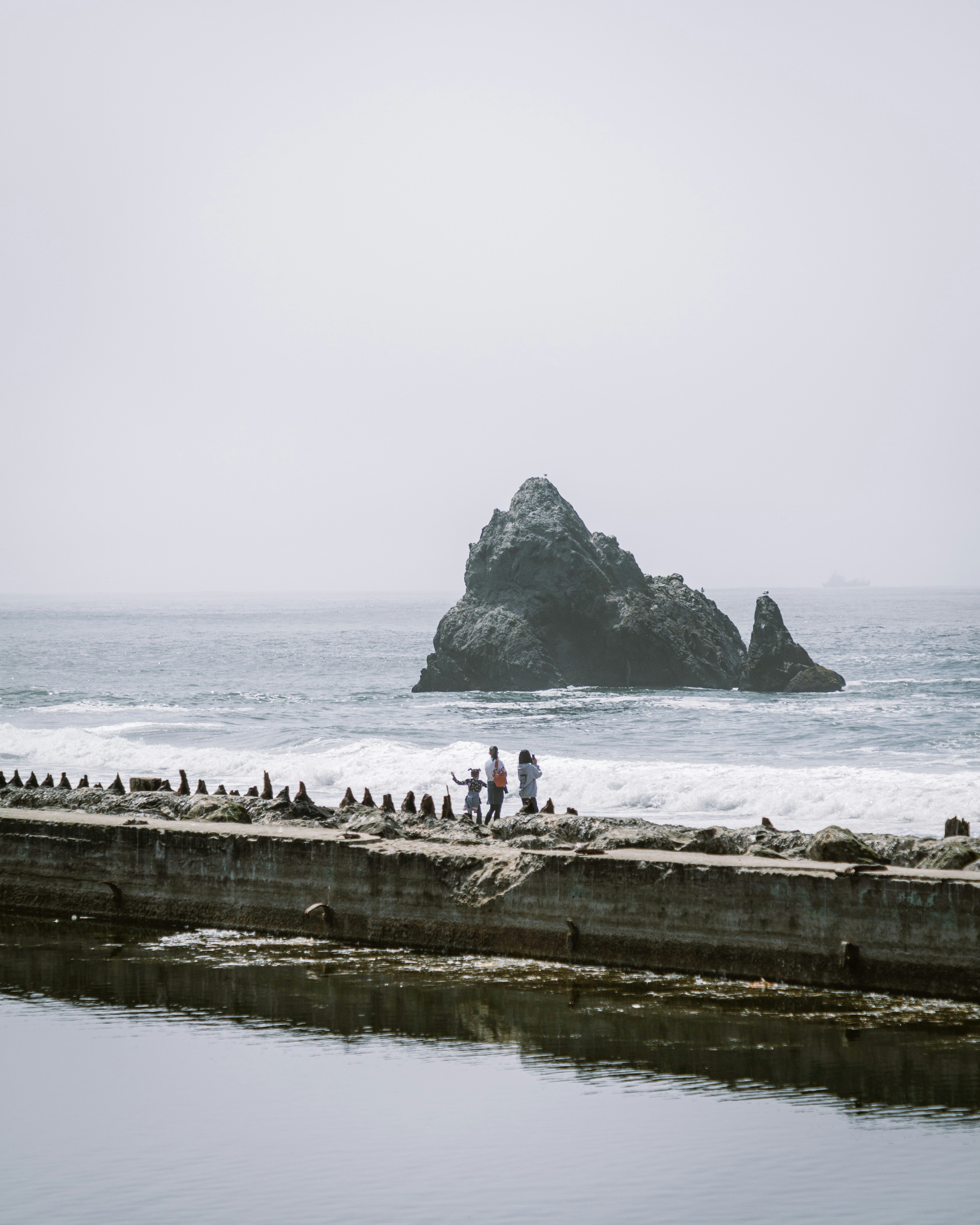 Family exploring the rocky shoreline near the ocean, with waves gently lapping at the beach. A prominent rock formation looms in the background.