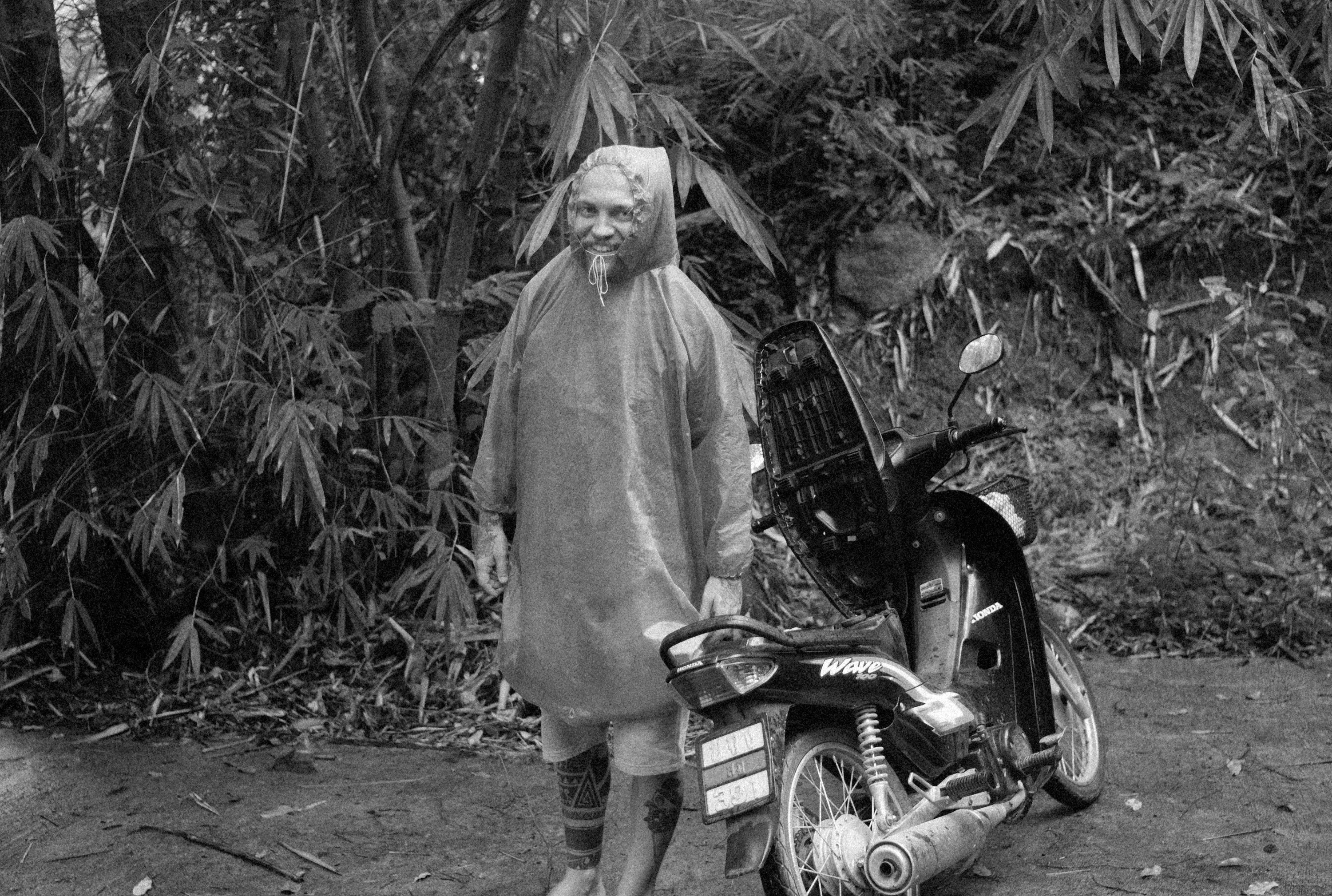 A figure in a raincoat stands beside a scooter, surrounded by dense bamboo foliage. The scene captures a moment of solitude in a lush, verdant environment.