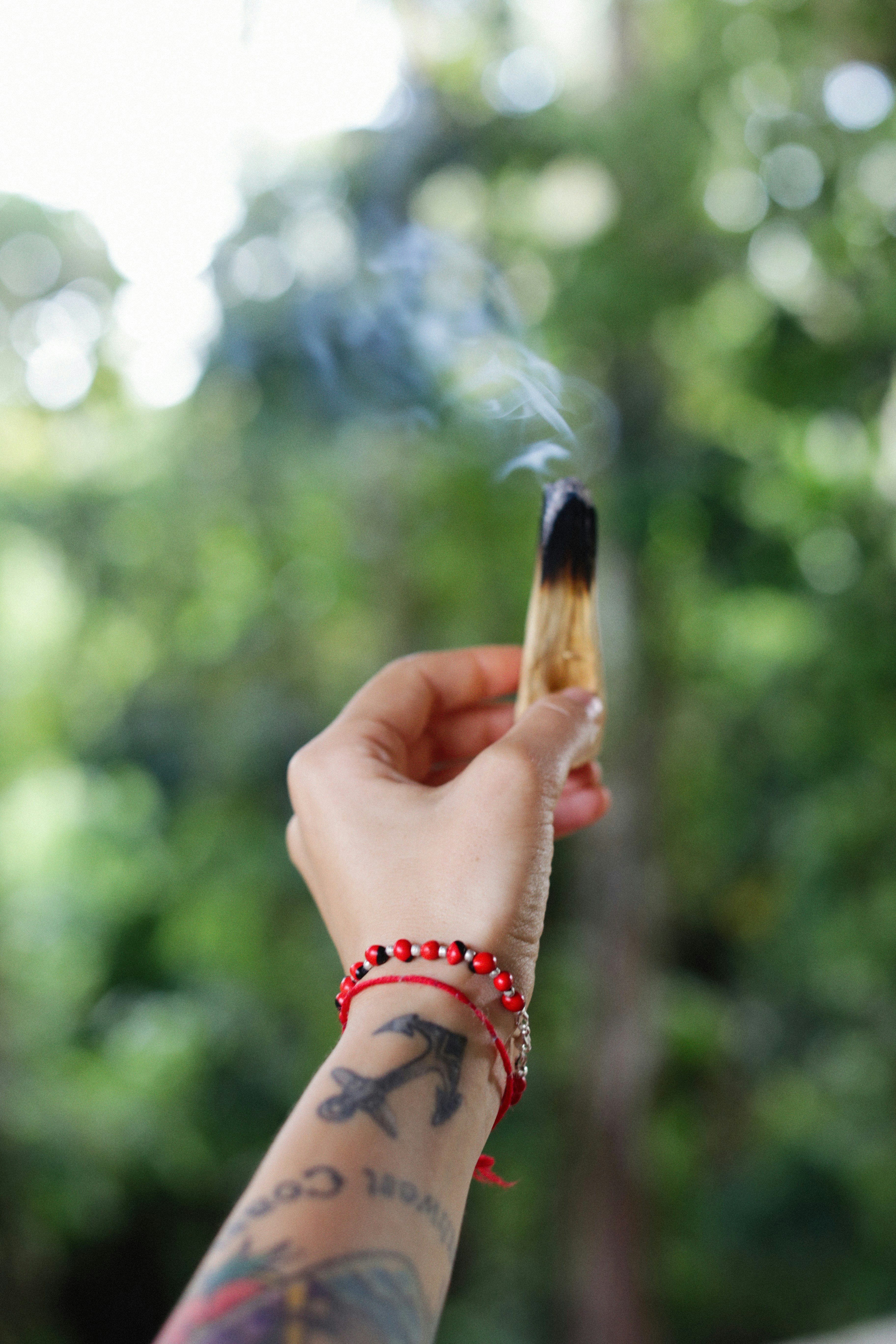 Hand holding a burning stick with smoke rising, set against a lush green backdrop. The image captures a moment of tranquility and connection with nature.