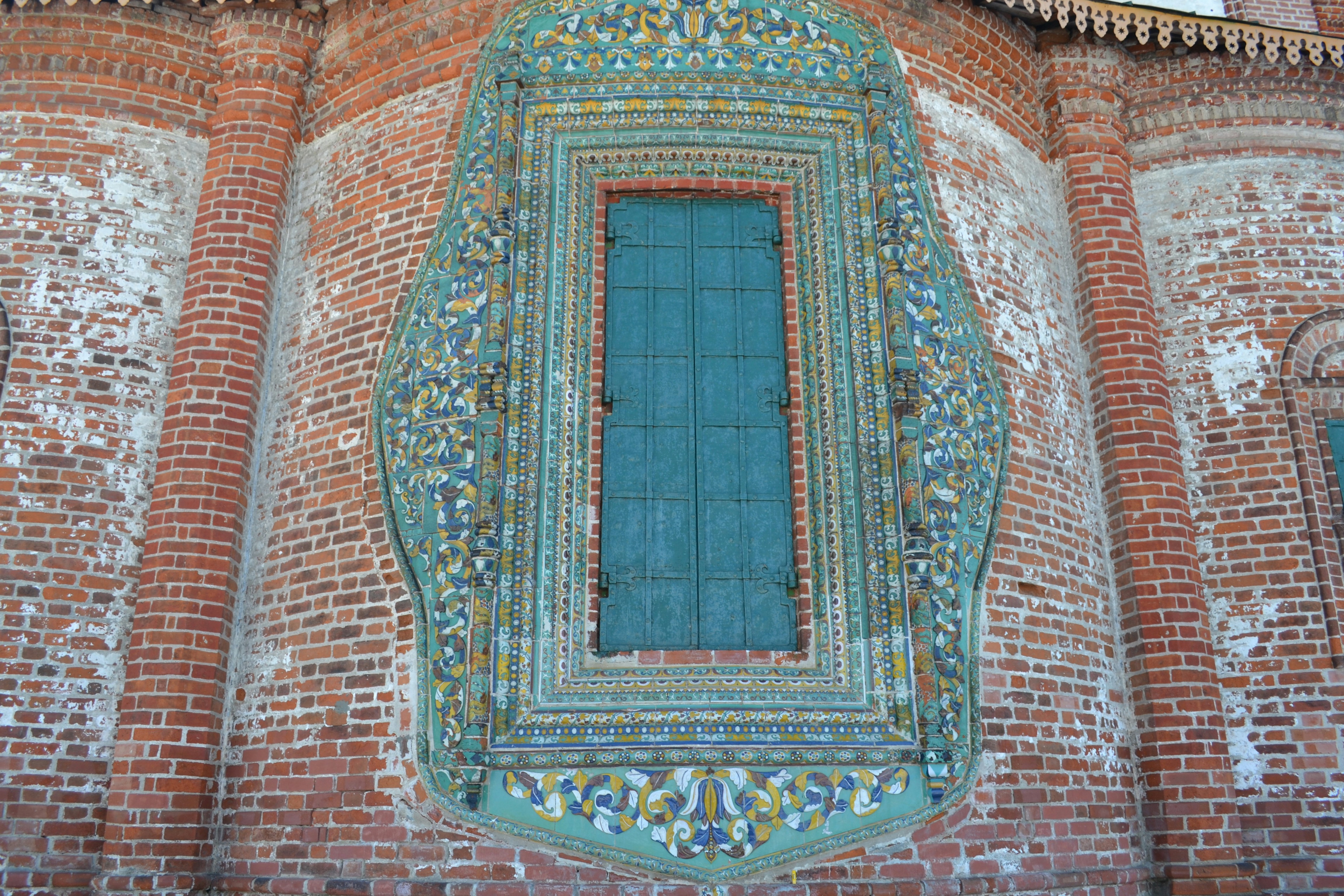 A vibrant mosaic frame surrounds a turquoise panel, set against the textured backdrop of aged red brick. The ornate design showcases intricate patterns and colors.
