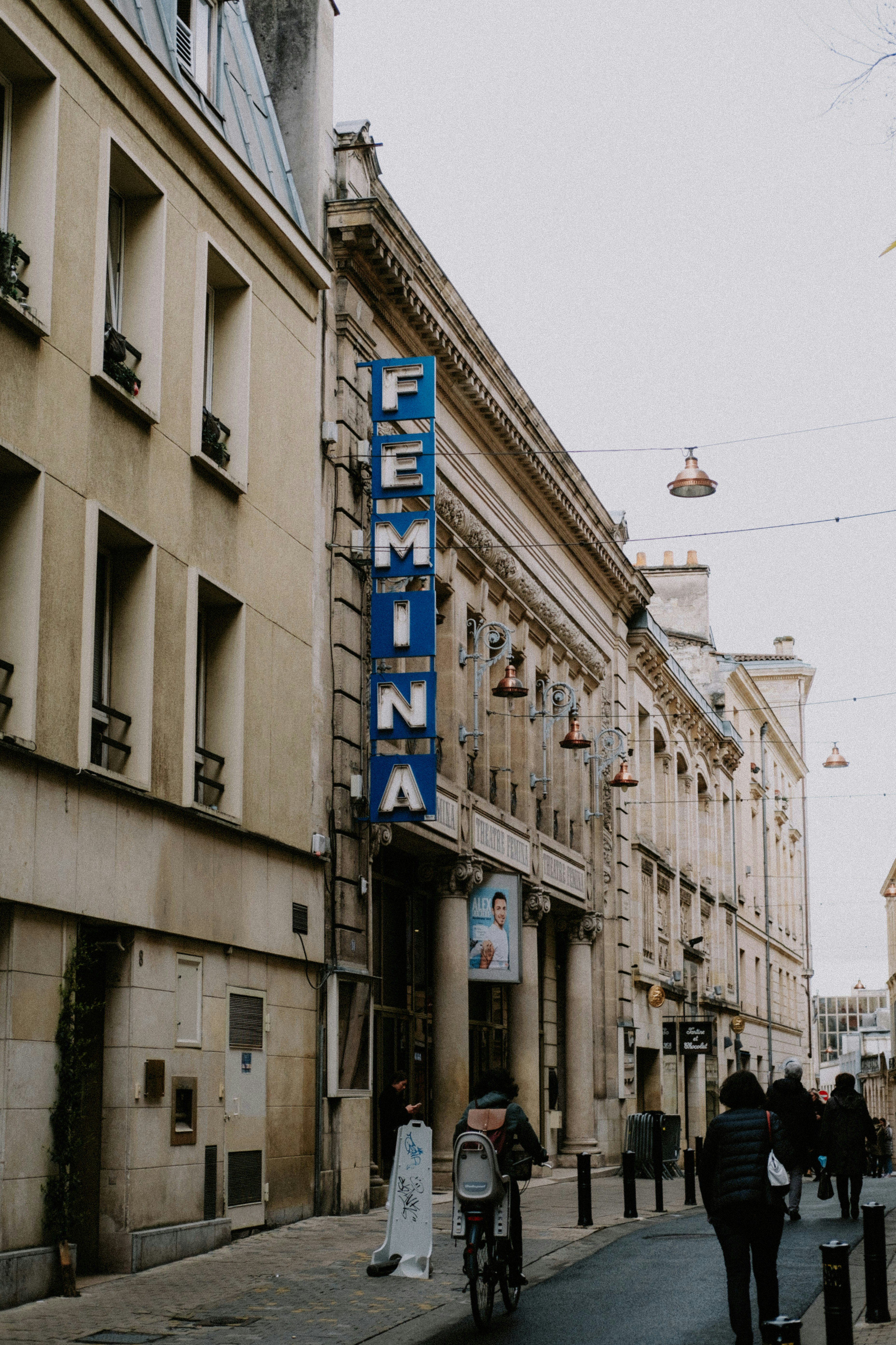 Historic Femina Theater sign illuminates a charming street, with pedestrians strolling past quaint architecture. 