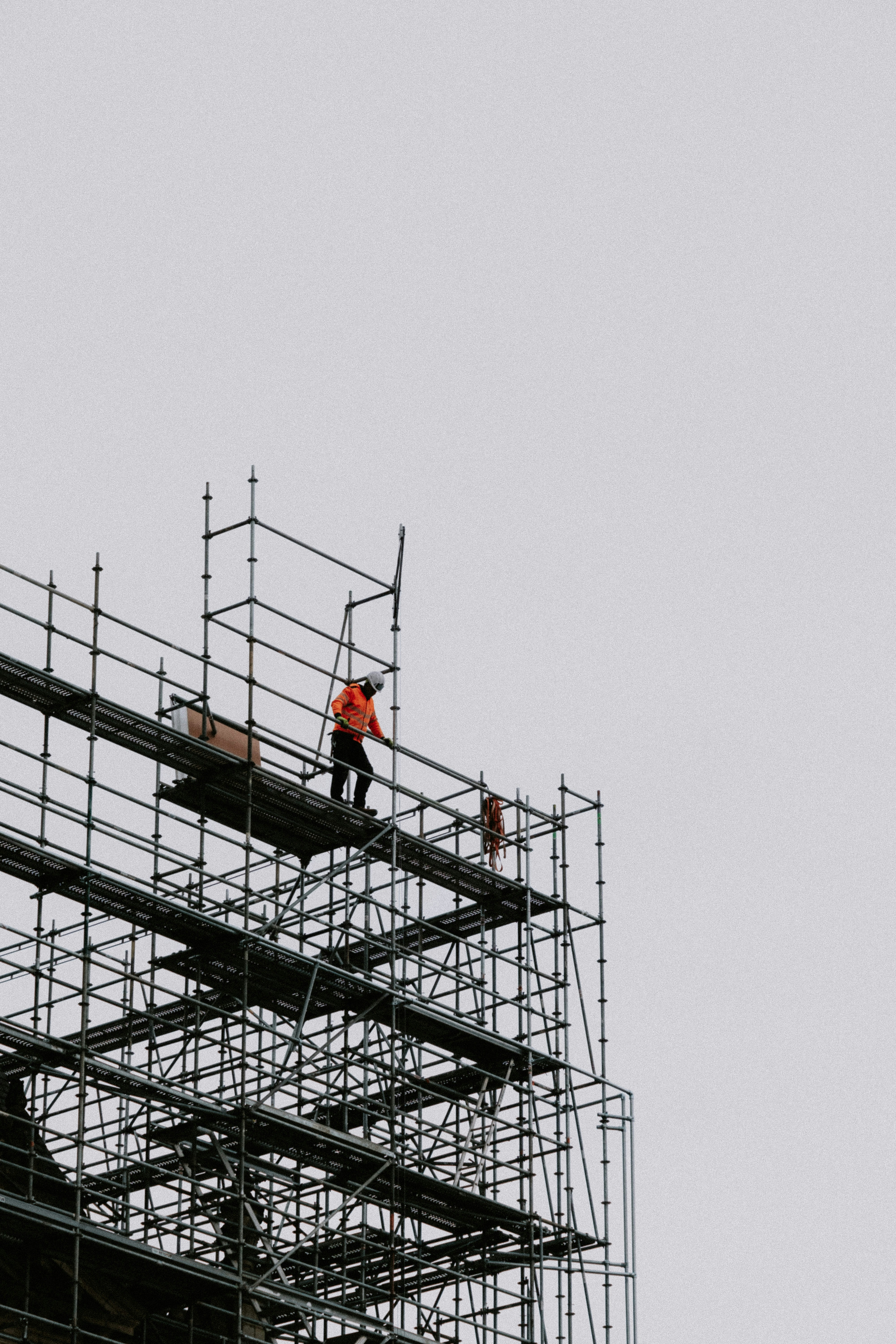 Construction worker navigating scaffolding on a building site against a muted sky.