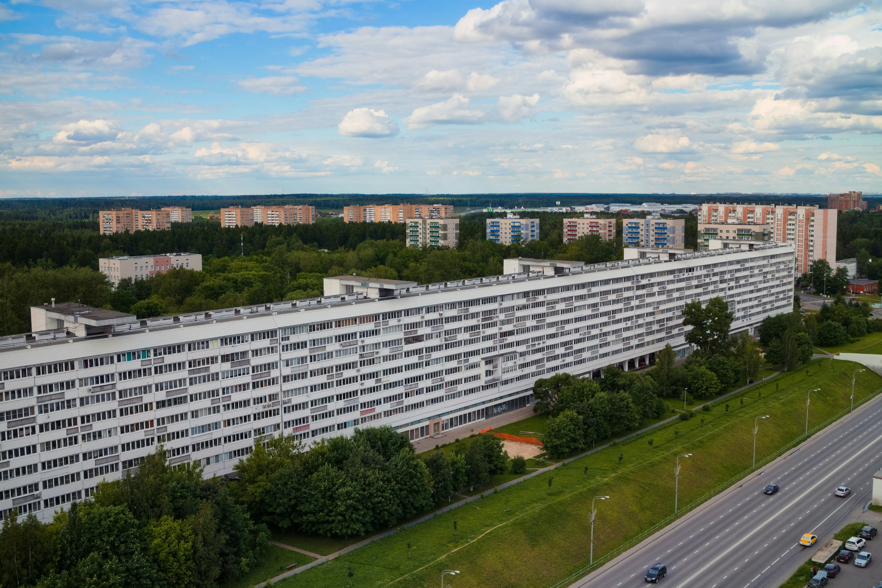 Long residential building stretches across a green landscape with scattered high-rise apartments under a partly cloudy sky.