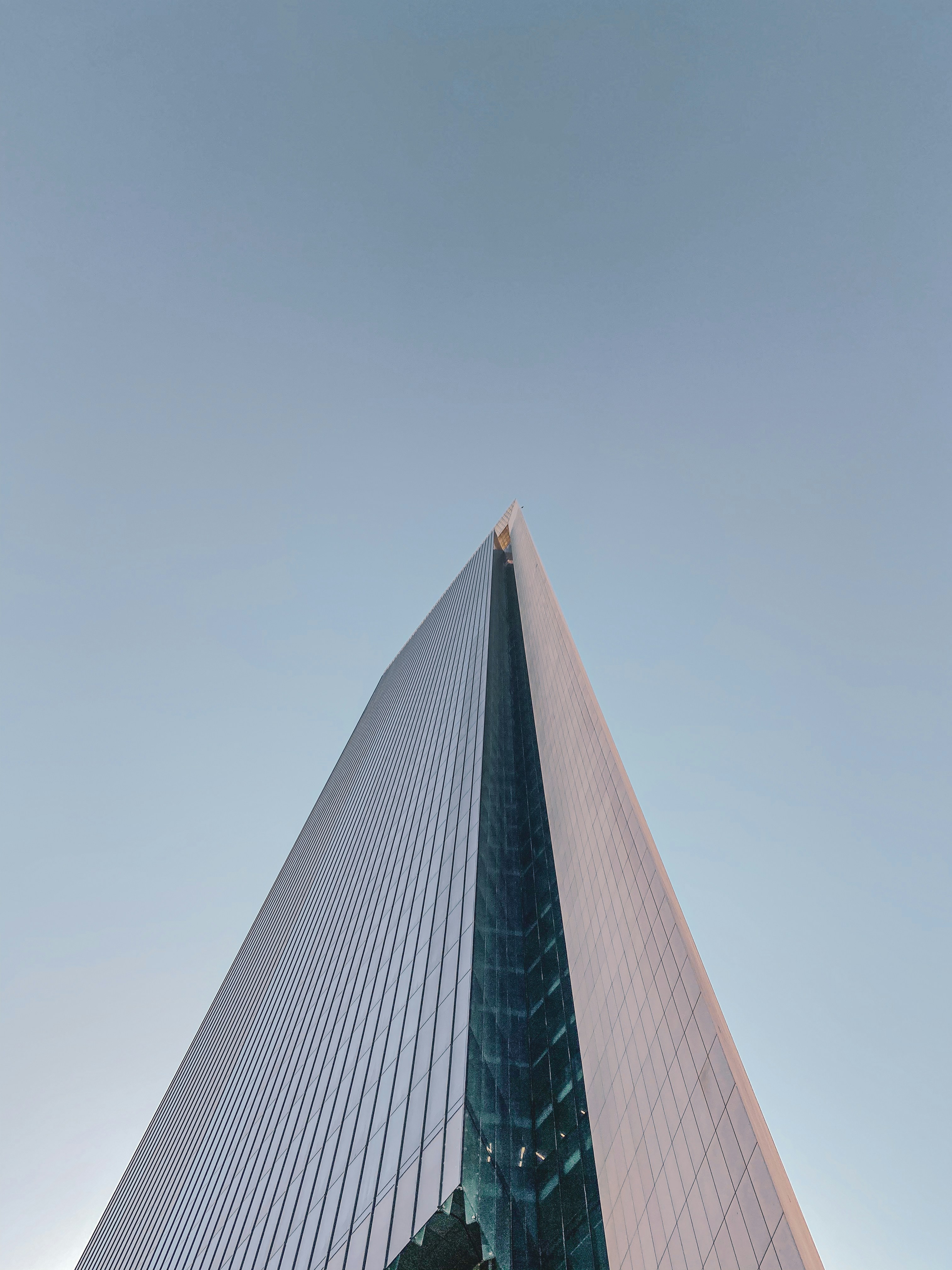 A towering skyscraper reaches towards the sky, showcasing its sleek glass facade against a pale blue backdrop.