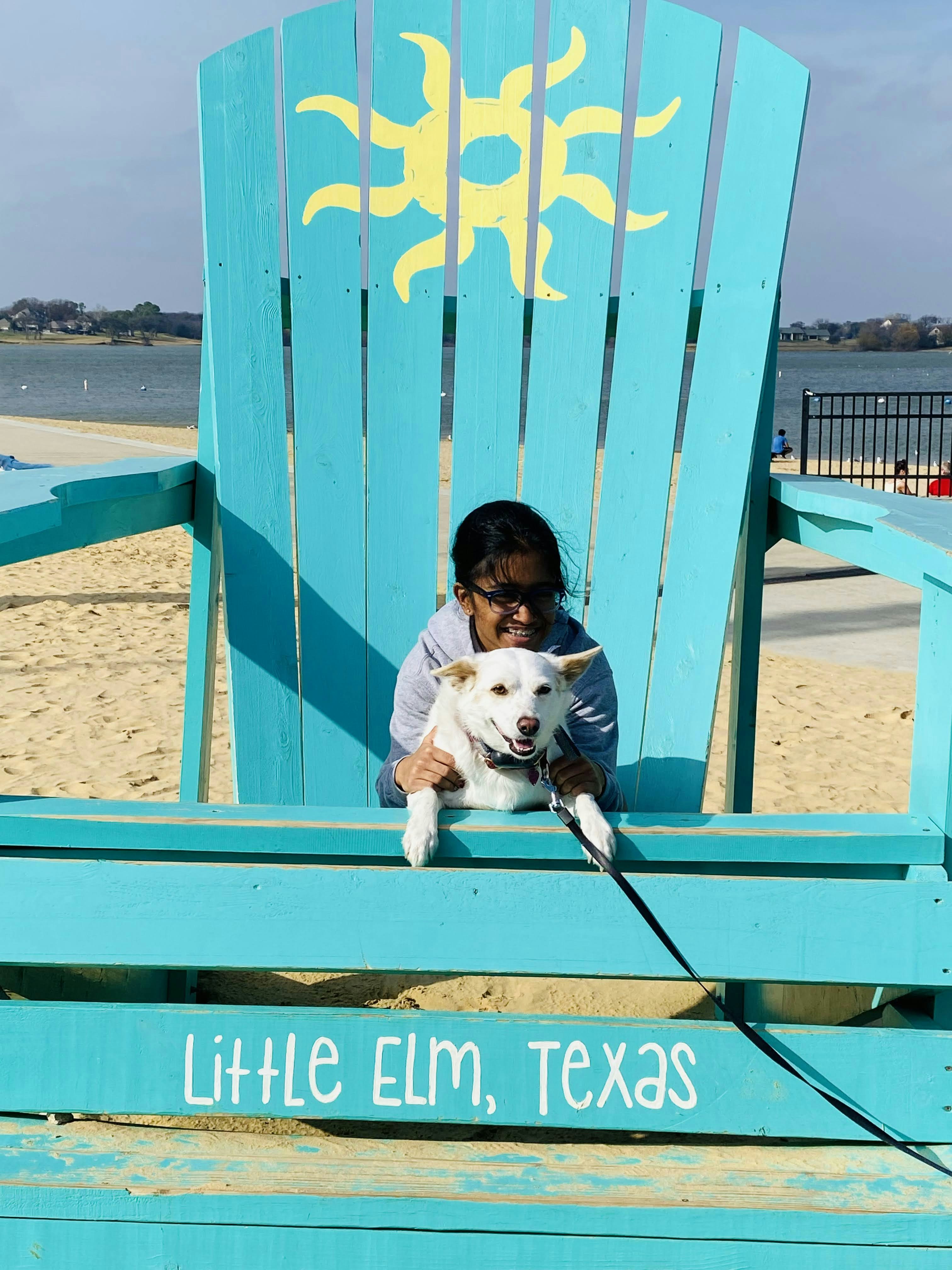 A girl smiles while holding her dog, sitting on a large turquoise chair adorned with a sun design, at a beach in Little Elm, Texas.
