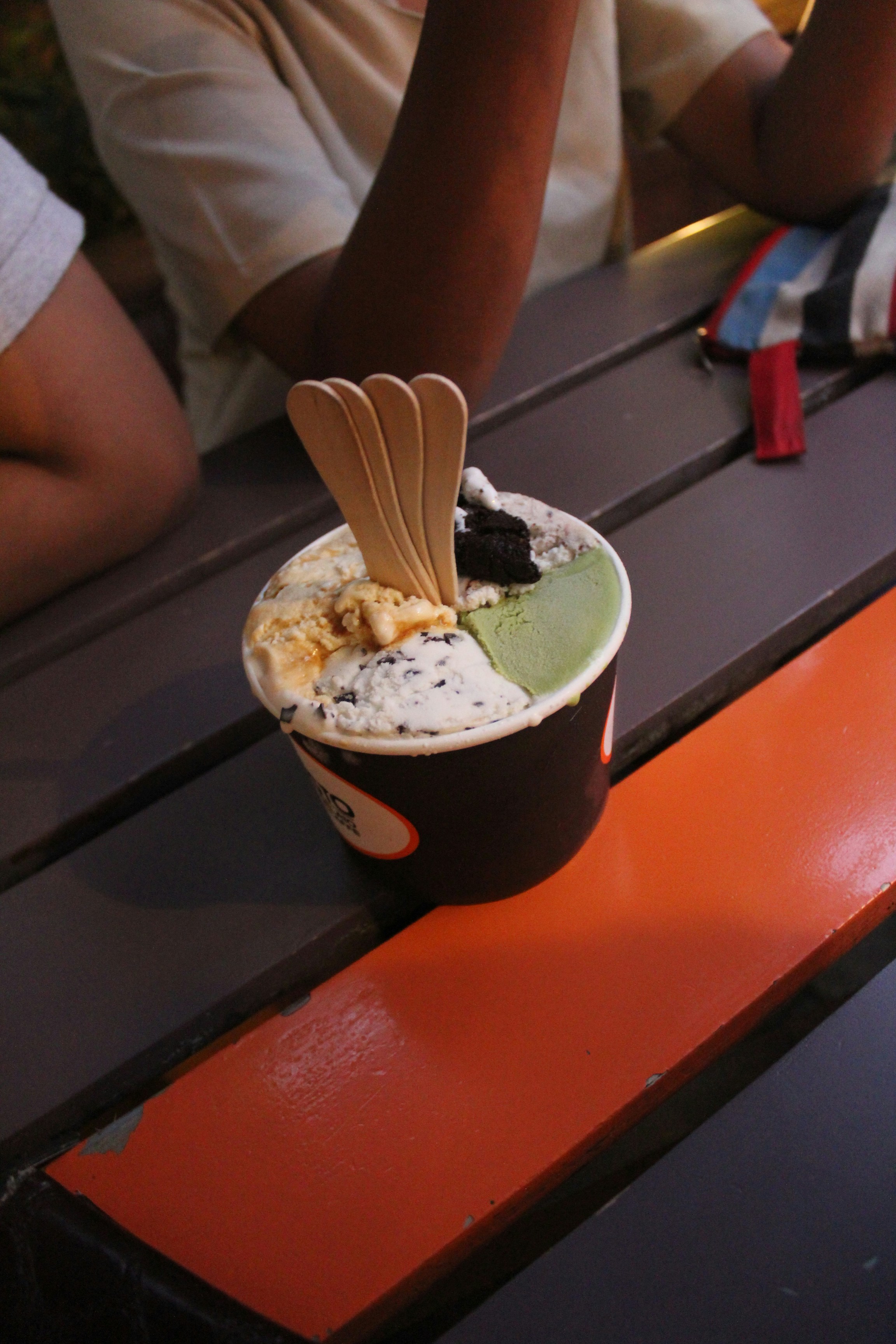 A colorful cup of ice cream featuring green tea, cookies and cream, and caramel flavors, with wooden spoons resting atop. The setting suggests a casual outdoor experience.