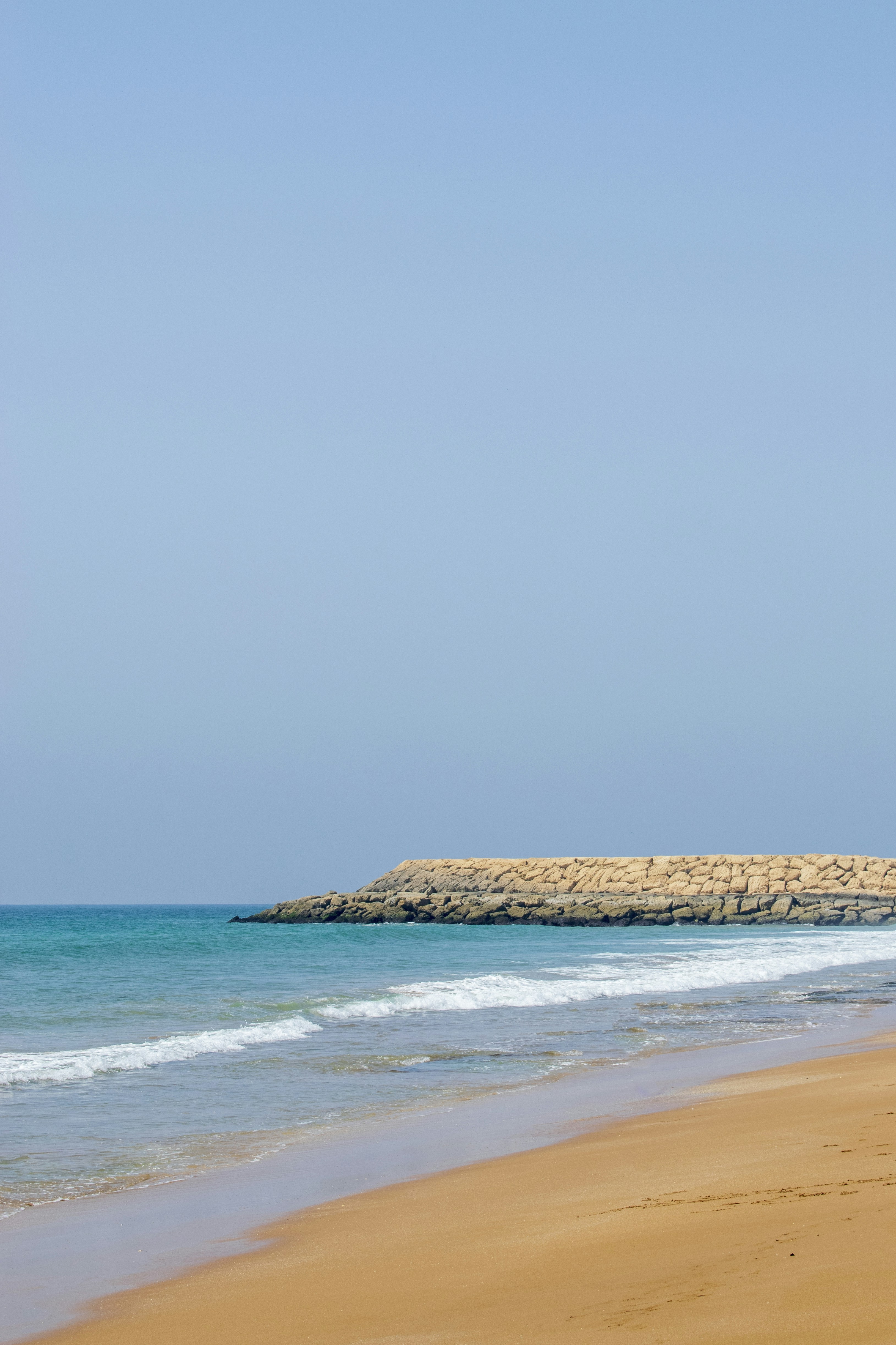 Calm beach scene featuring gentle waves lapping against a stone jetty under a clear blue sky.