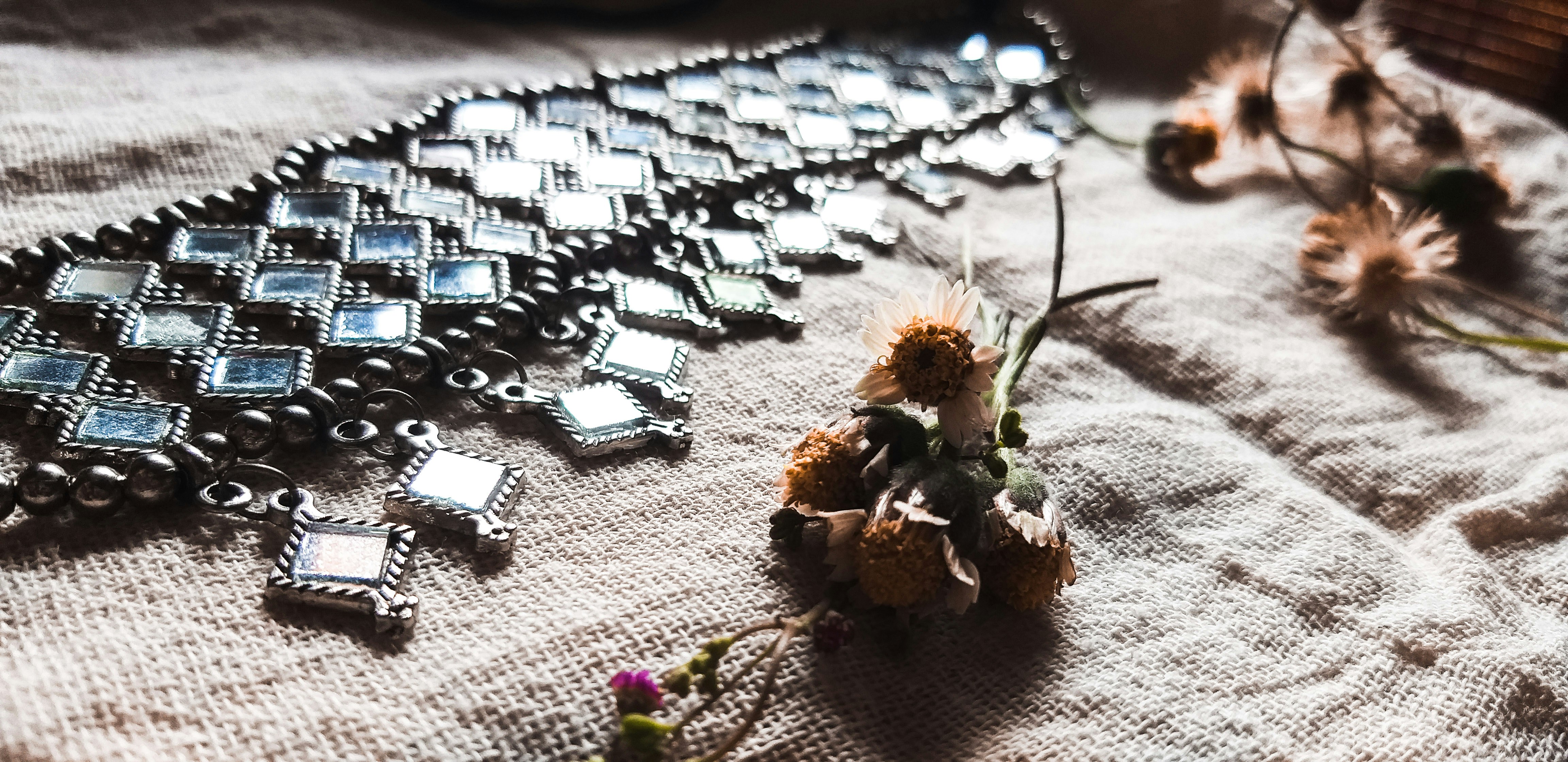 Intricate silver jewelry piece showcased alongside dried flowers on a textured fabric background.