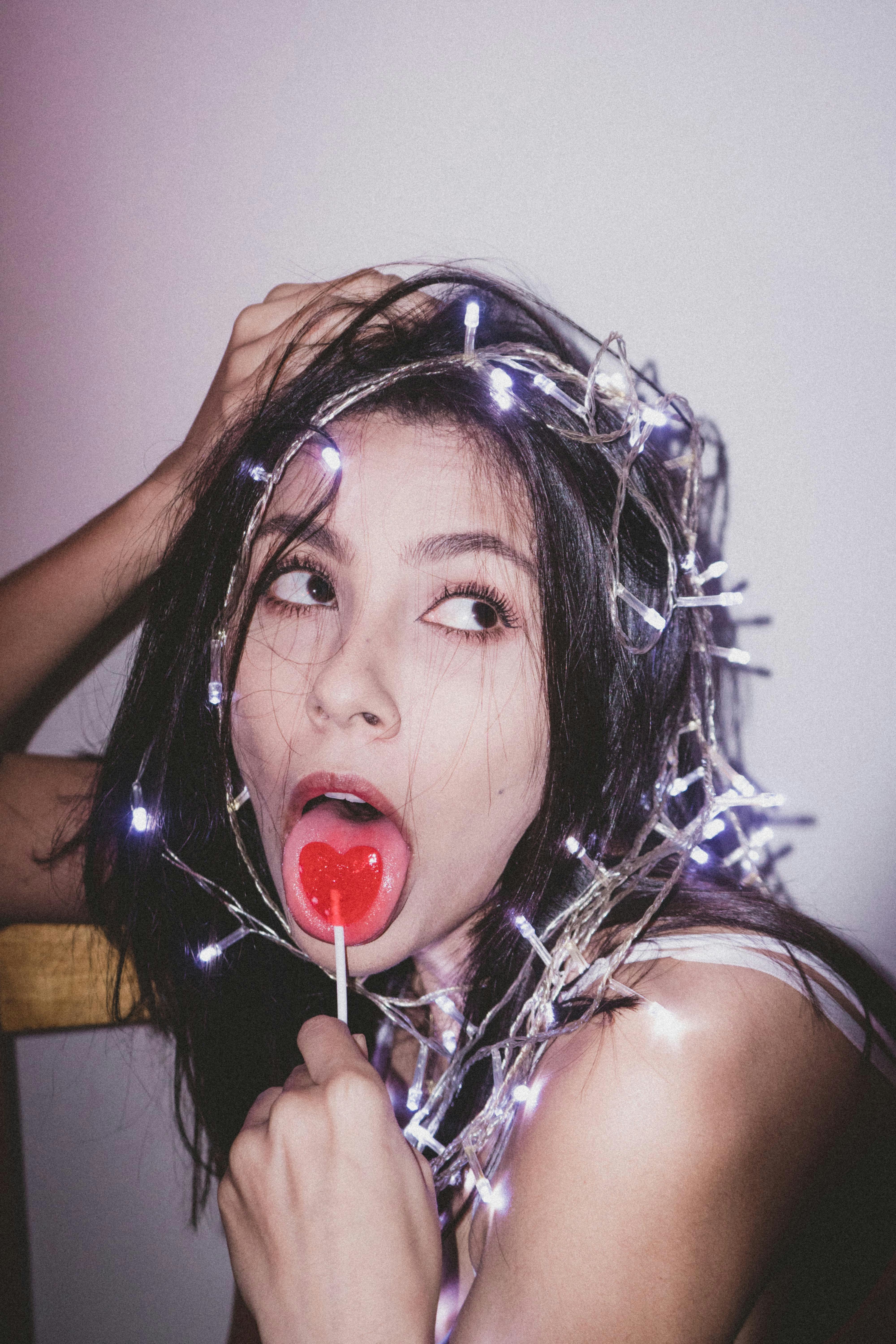 A young woman playfully poses with a heart-shaped lollipop, her hair adorned with twinkling fairy lights against a soft backdrop.