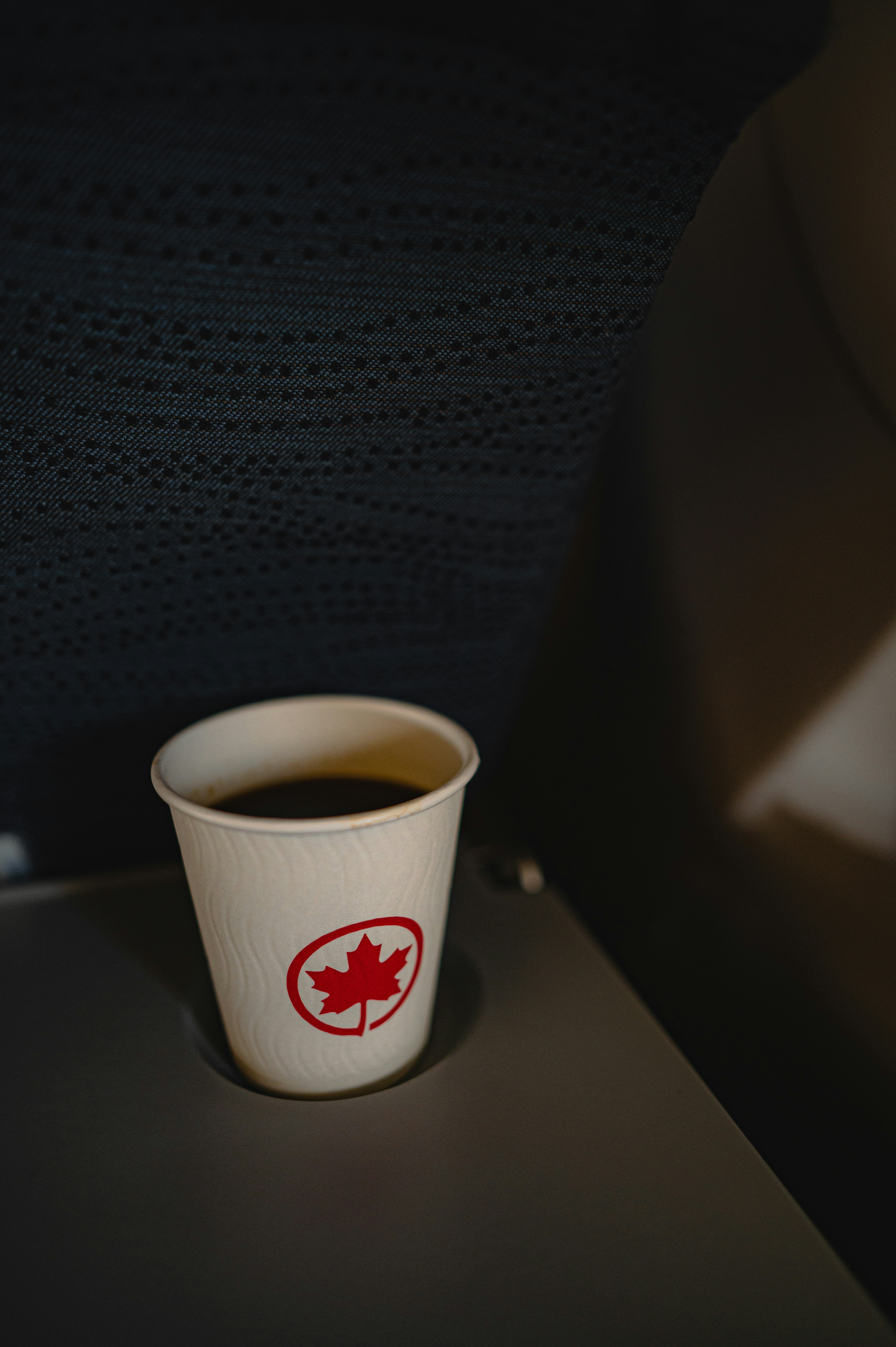 Coffee cup with a red maple leaf logo resting on an airplane tray table, surrounded by a textured fabric background.