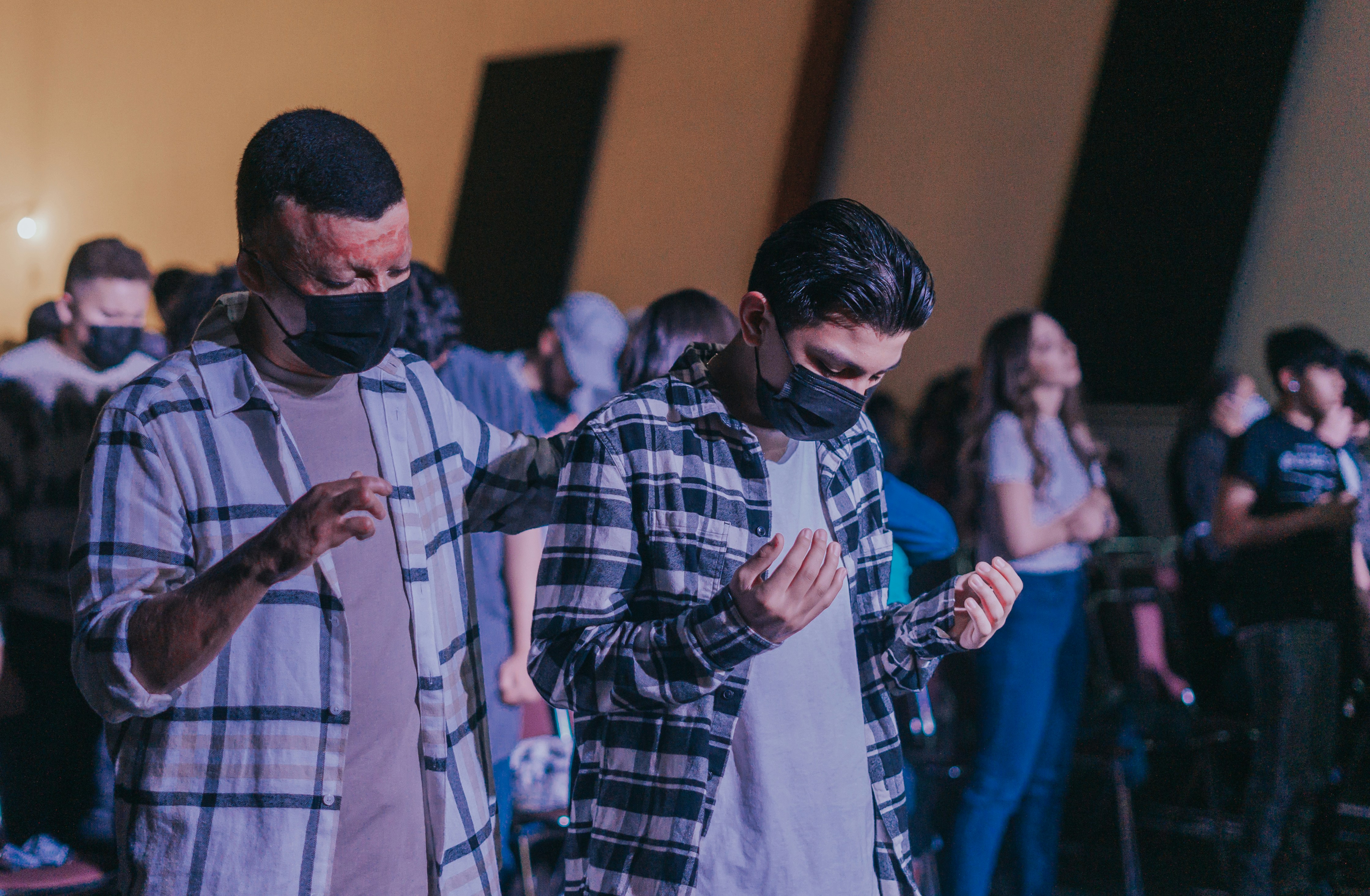 Two individuals in plaid shirts and masks share a moment of introspection amidst a gathering, surrounded by a crowd engaged in a shared experience.