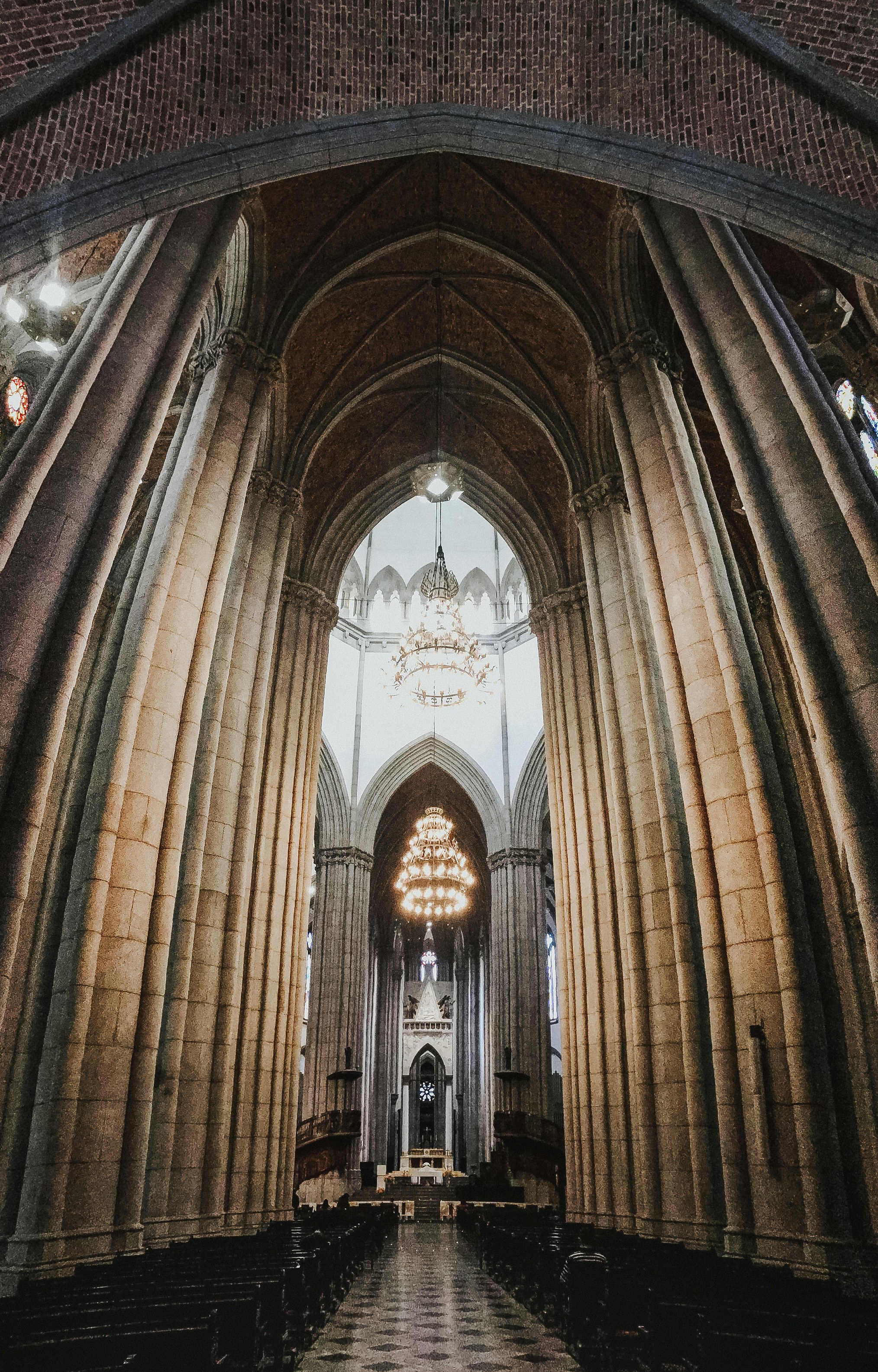 Interior of a grand cathedral showcasing soaring arches and intricate chandeliers. The light filters through stained glass, illuminating the space.