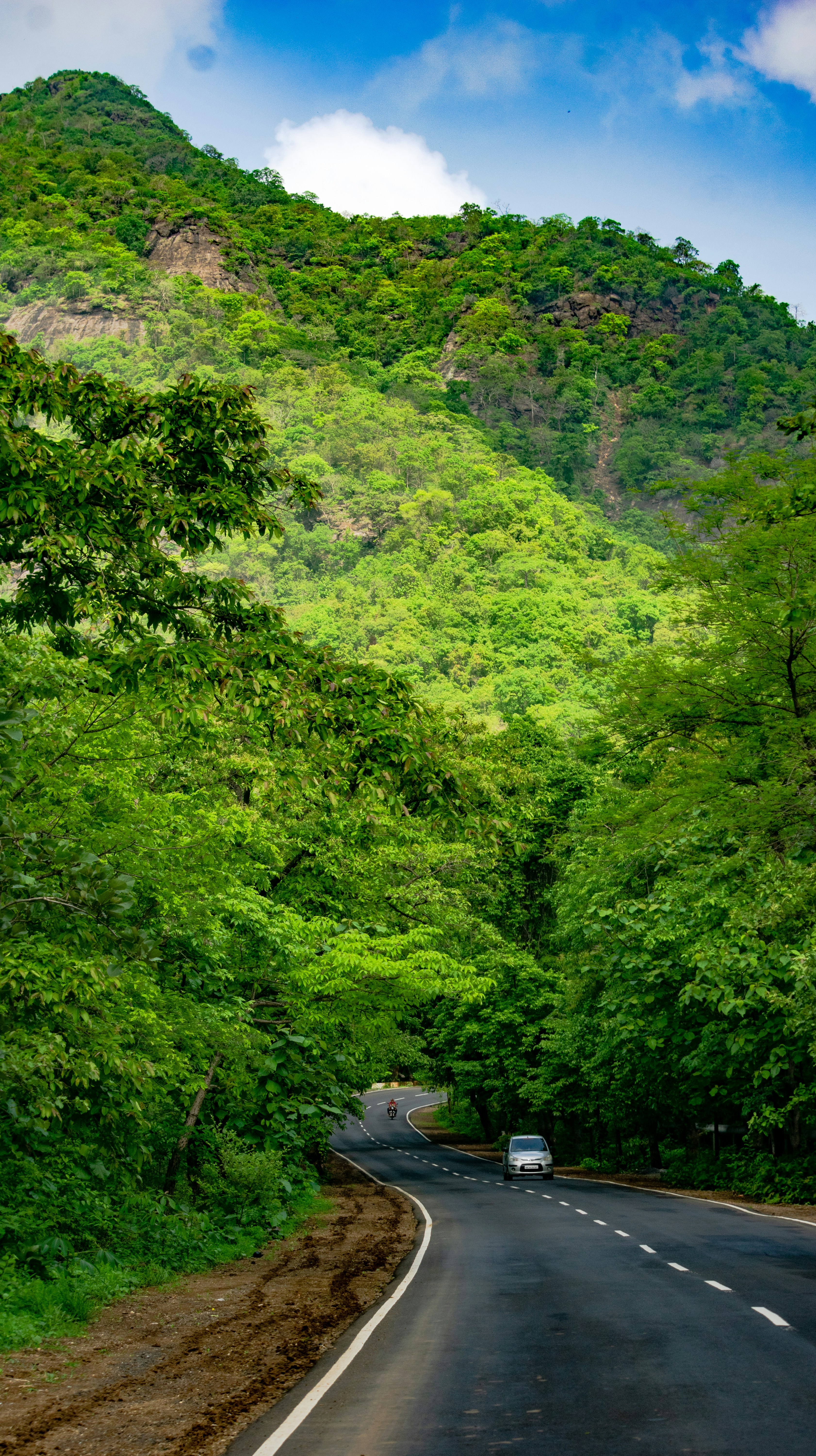 Curved road flanked by lush greenery and hills under a partly cloudy sky.