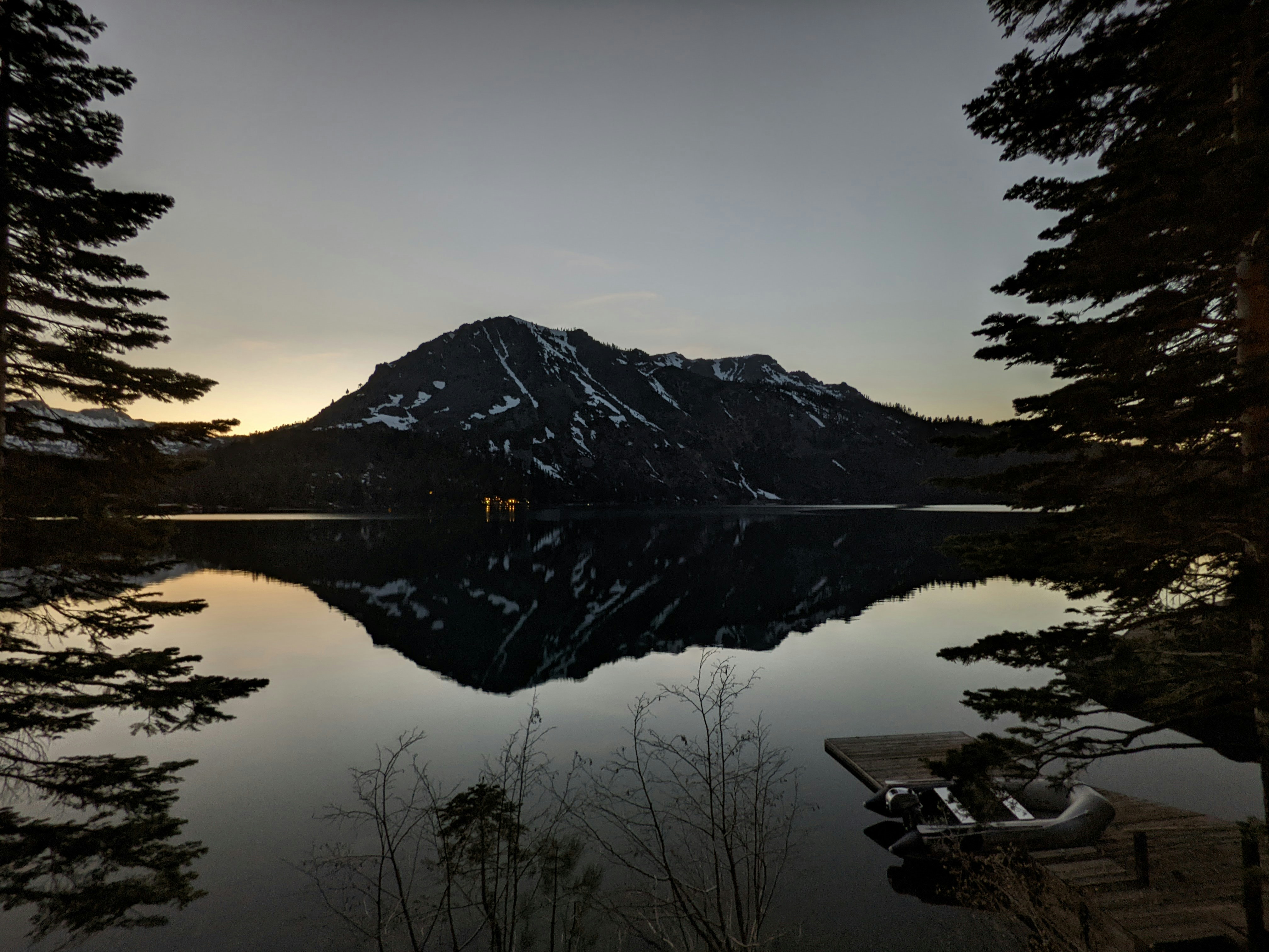 Serene lake reflecting a mountain silhouette under a twilight sky, framed by trees on either side. The stillness of the water enhances the tranquil atmosphere.