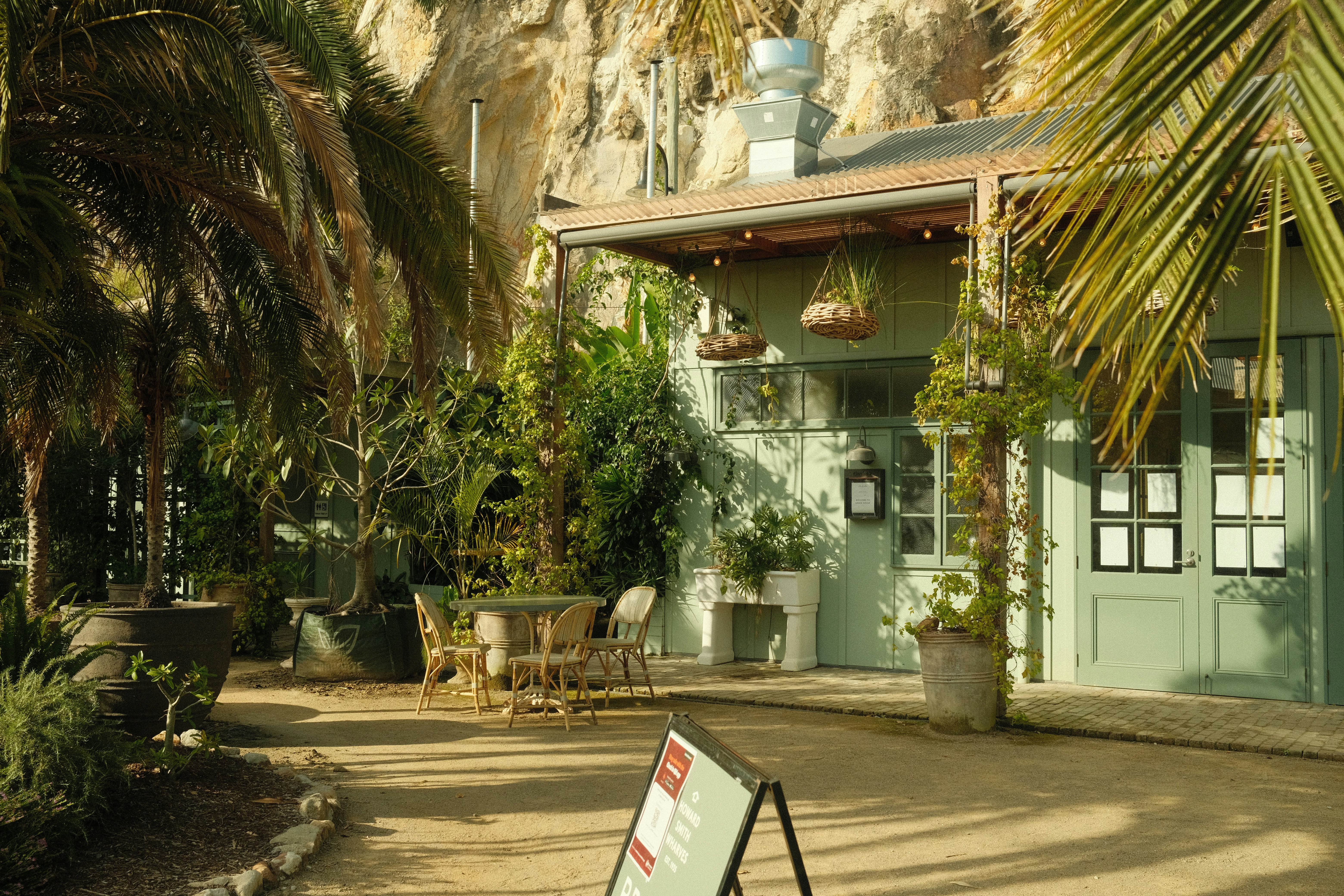 Outdoor cafe with green facade surrounded by lush plants and palm trees under warm sunlight.