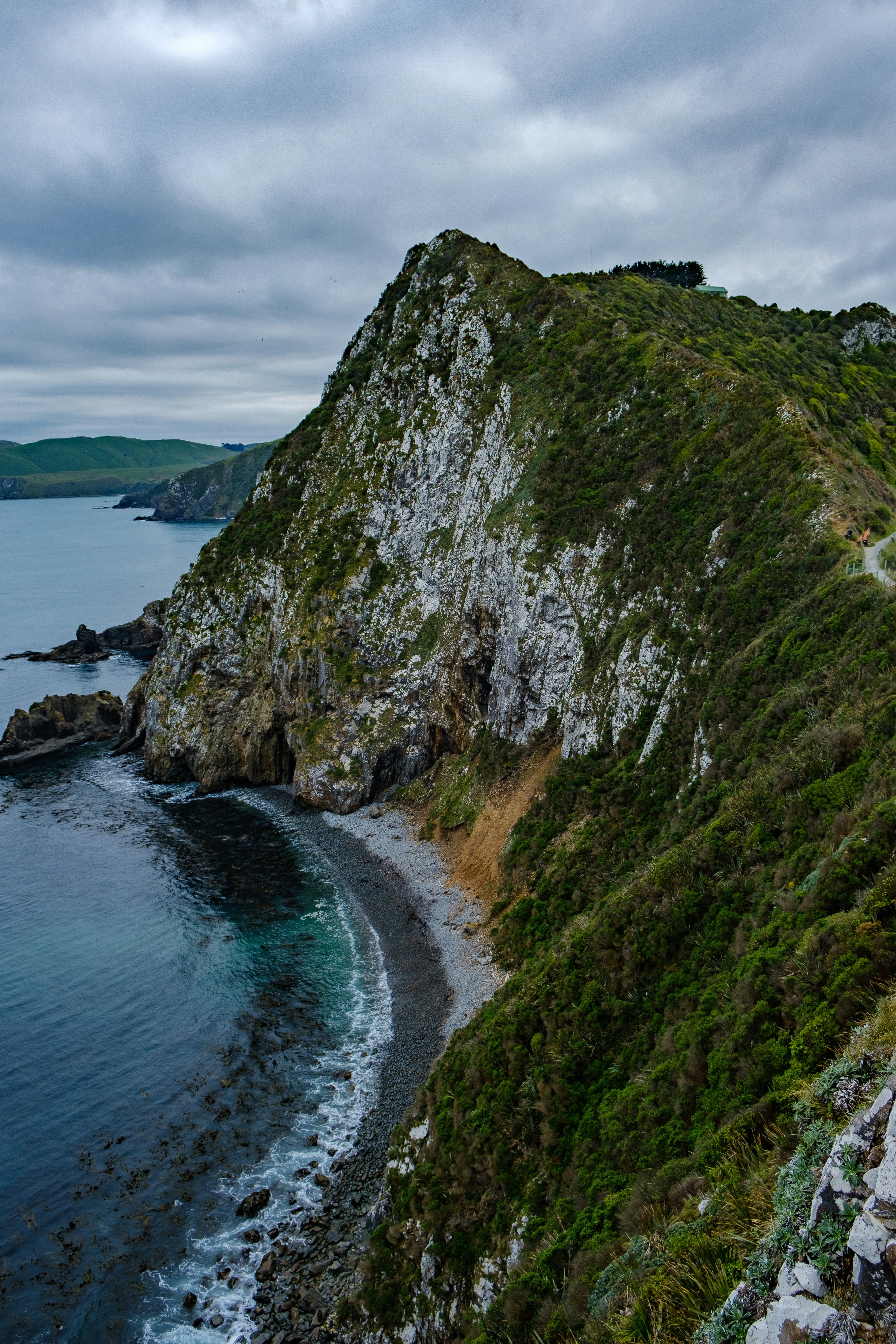 Dramatic coastal cliffs rise above a tranquil bay, where waves gently lap at a pebbled shore. The lush greenery contrasts with the rugged rock face.