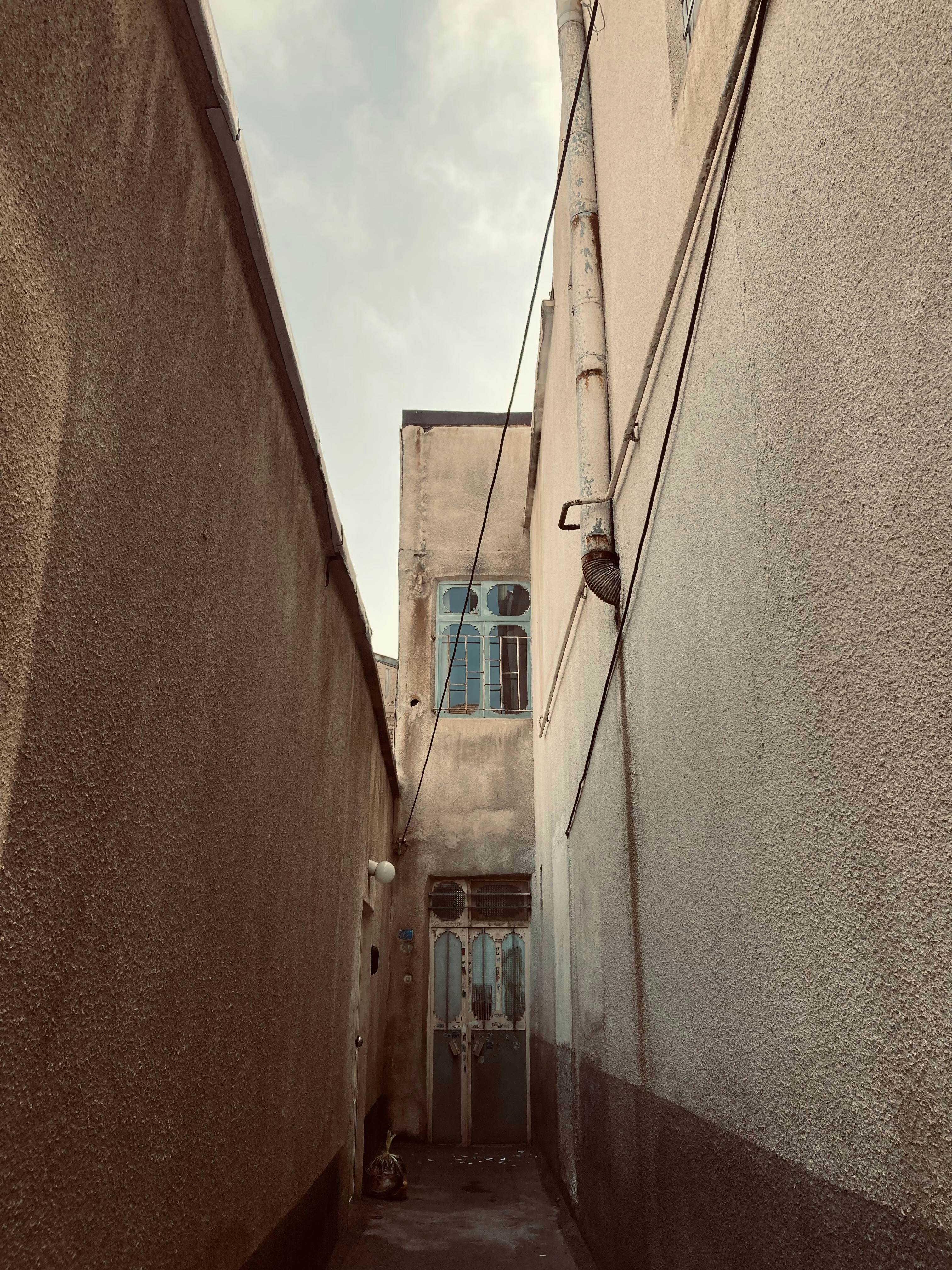 Narrow alleyway flanked by textured walls, leading to a vintage door with a blue window above. Power lines stretch overhead, adding to the scene's character.