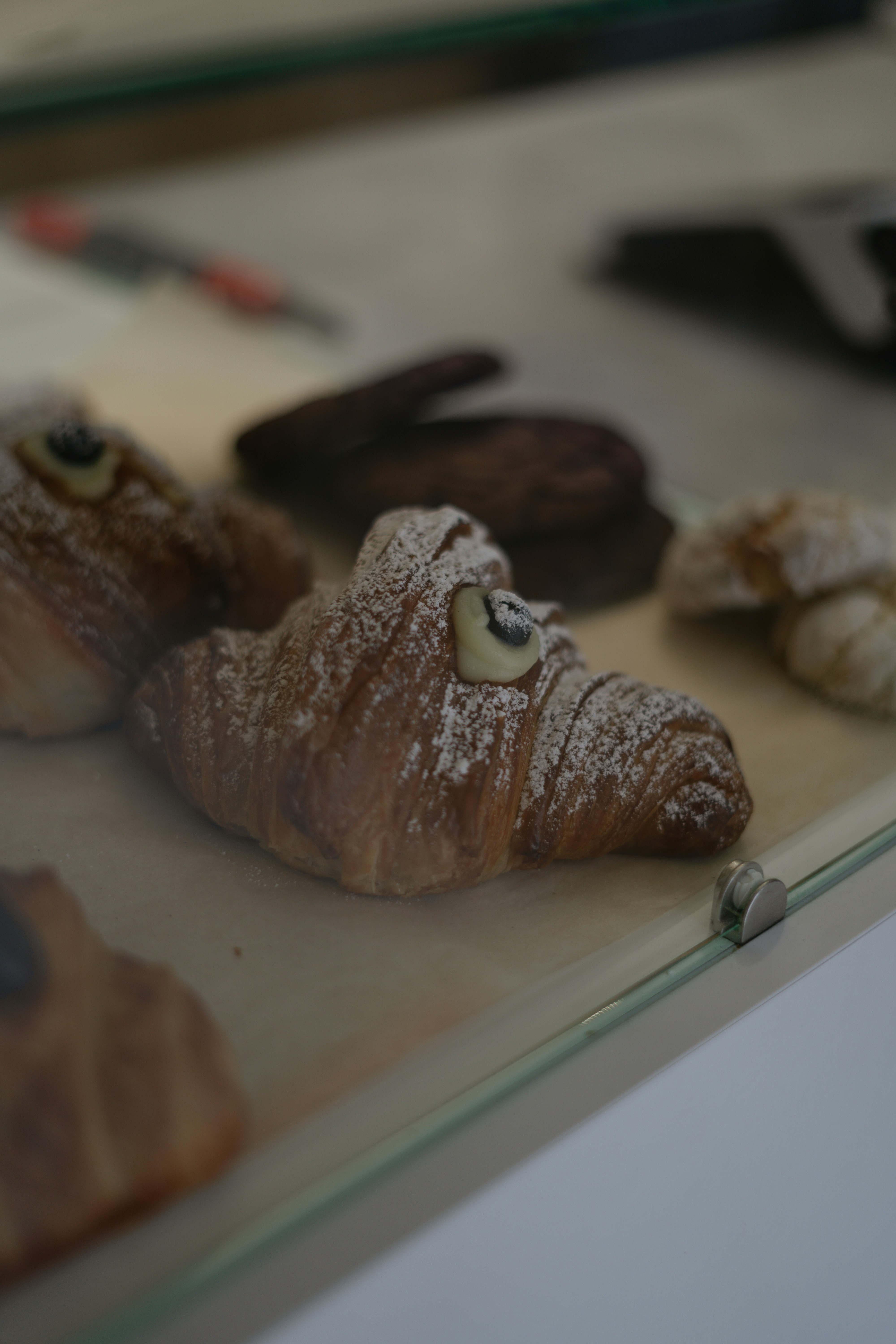 Crusty croissants with almond-like cream centers sit under glass in a bakery display case, dusted with powdered sugar. A warm, inviting pastry arrangement.