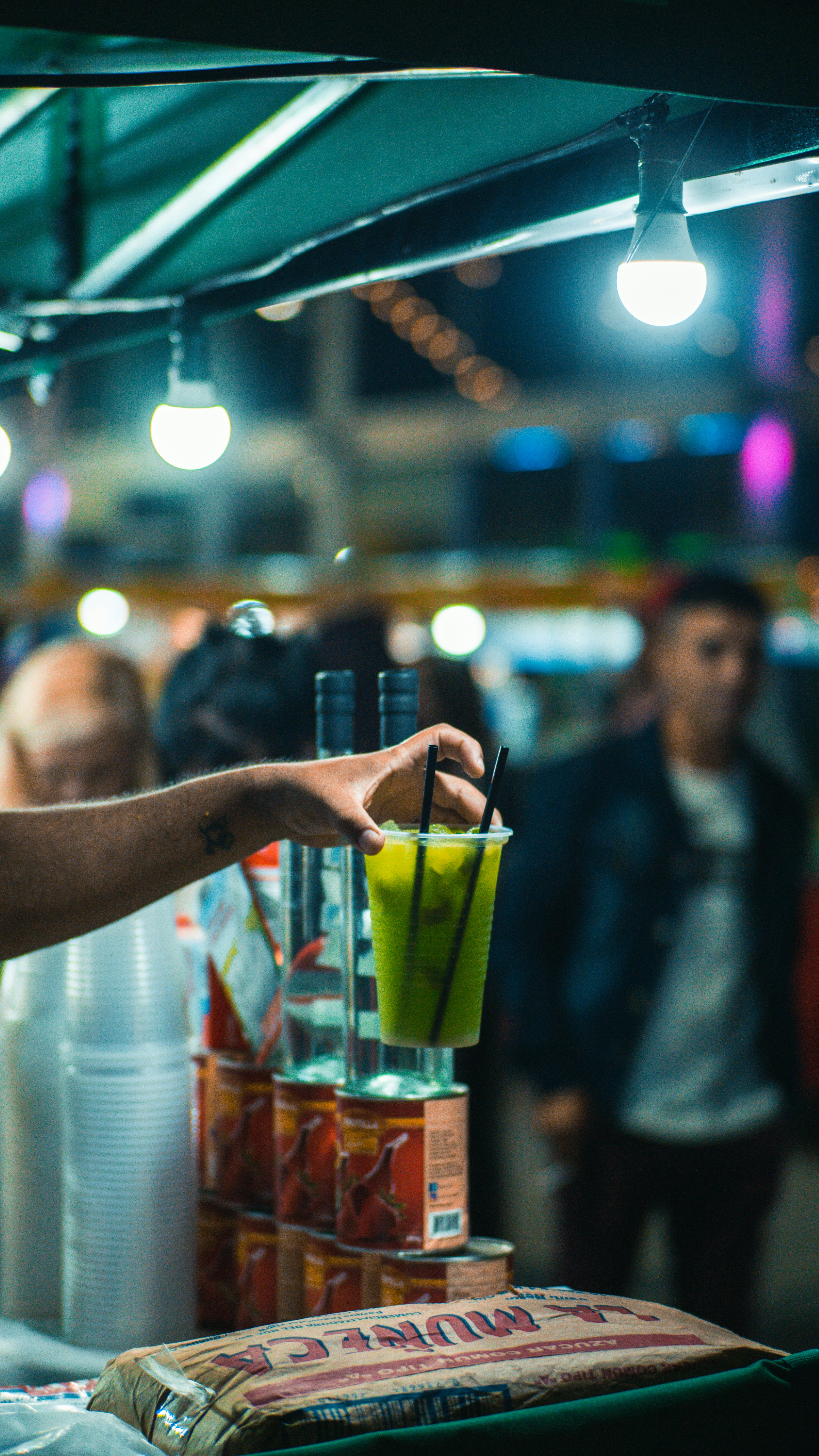 Hand holding a vibrant green drink with two straws against a bustling night market backdrop.