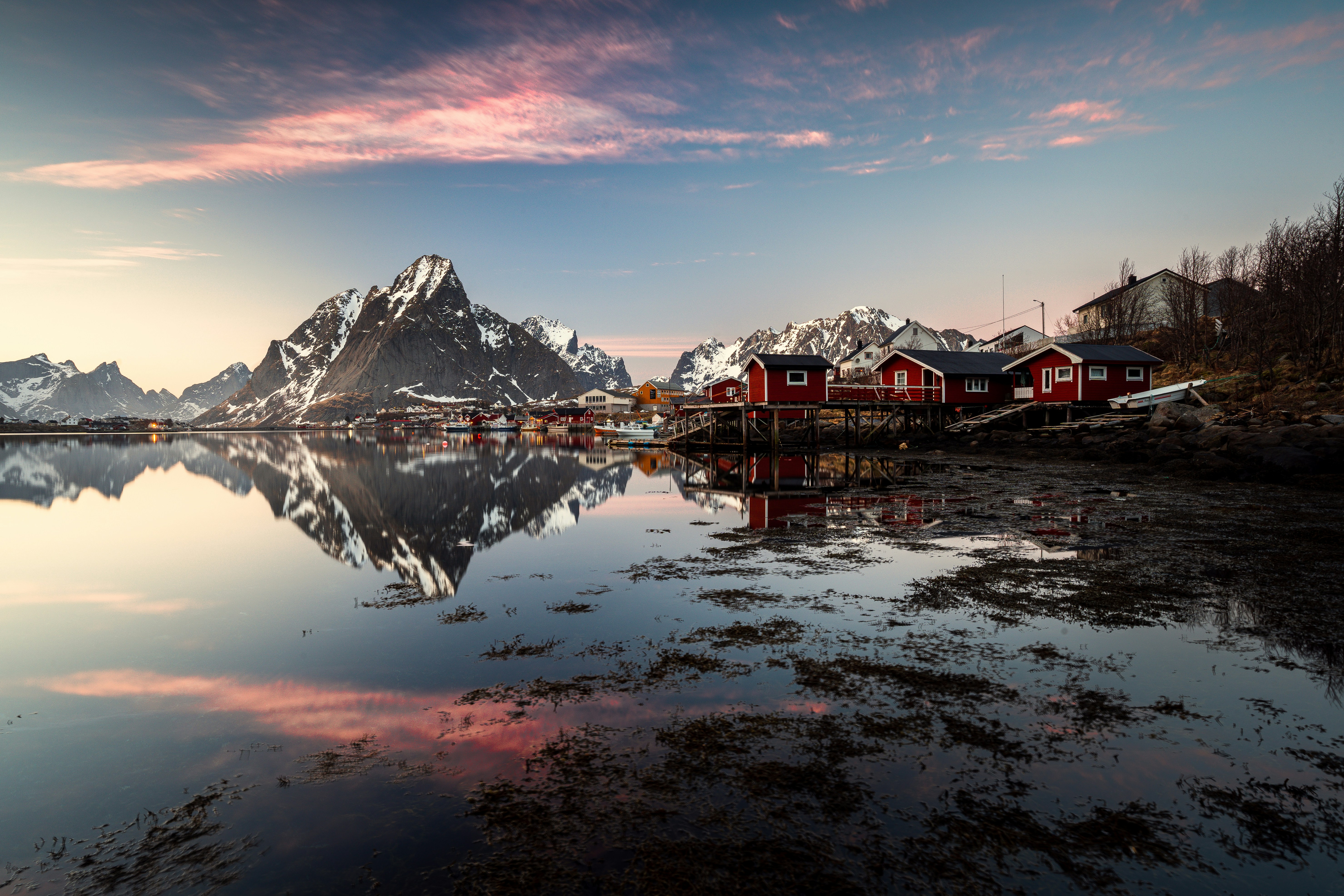 Snow-capped mountains and red cabins mirrored in calm waters under a pastel sky.