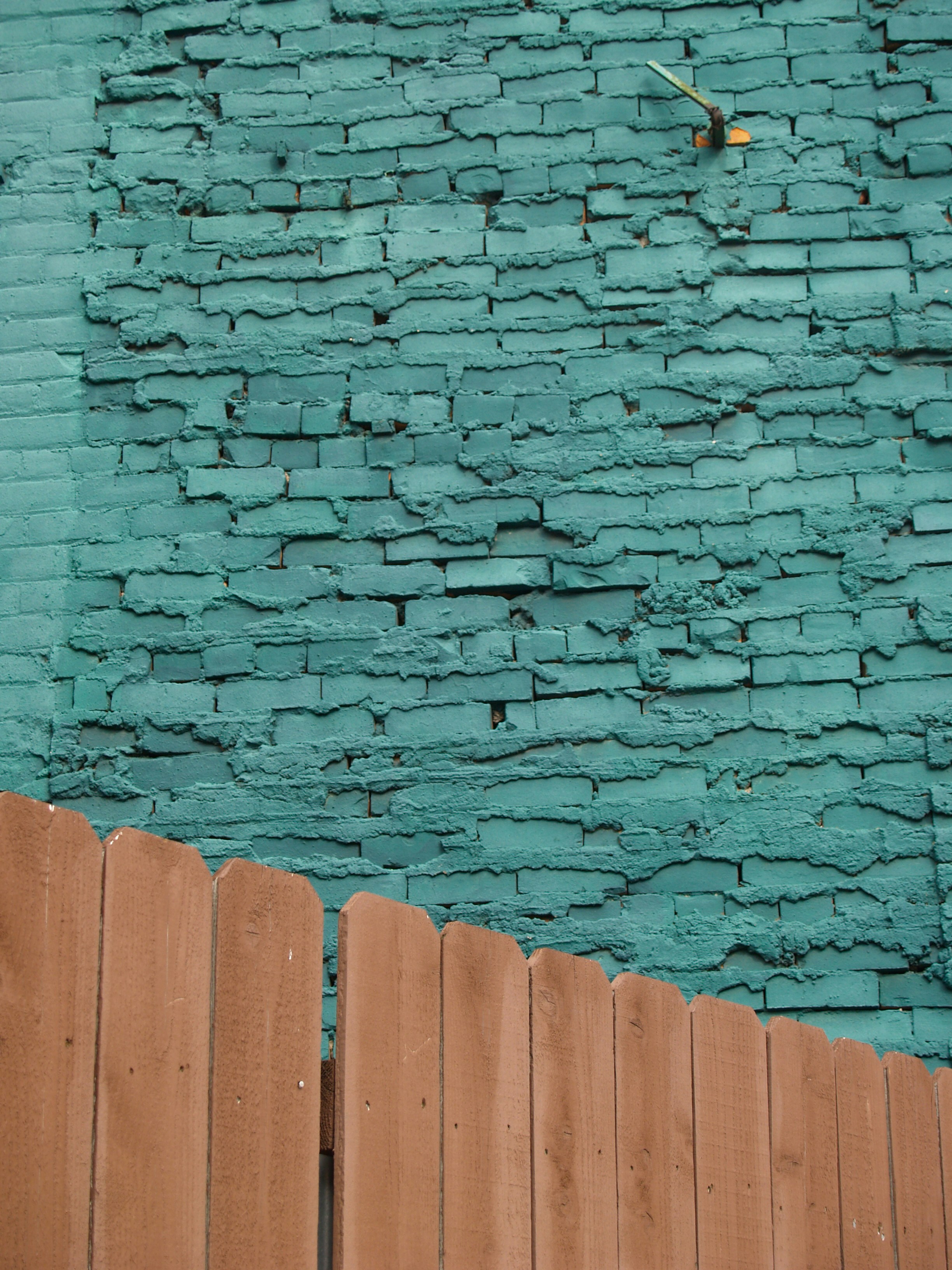 Weathered teal plaster wall with deep, irregular cracks dominates the frame, complemented by a row of vertical wooden fence slats in the foreground. A small orange detail near the top provides a subtle focal point.