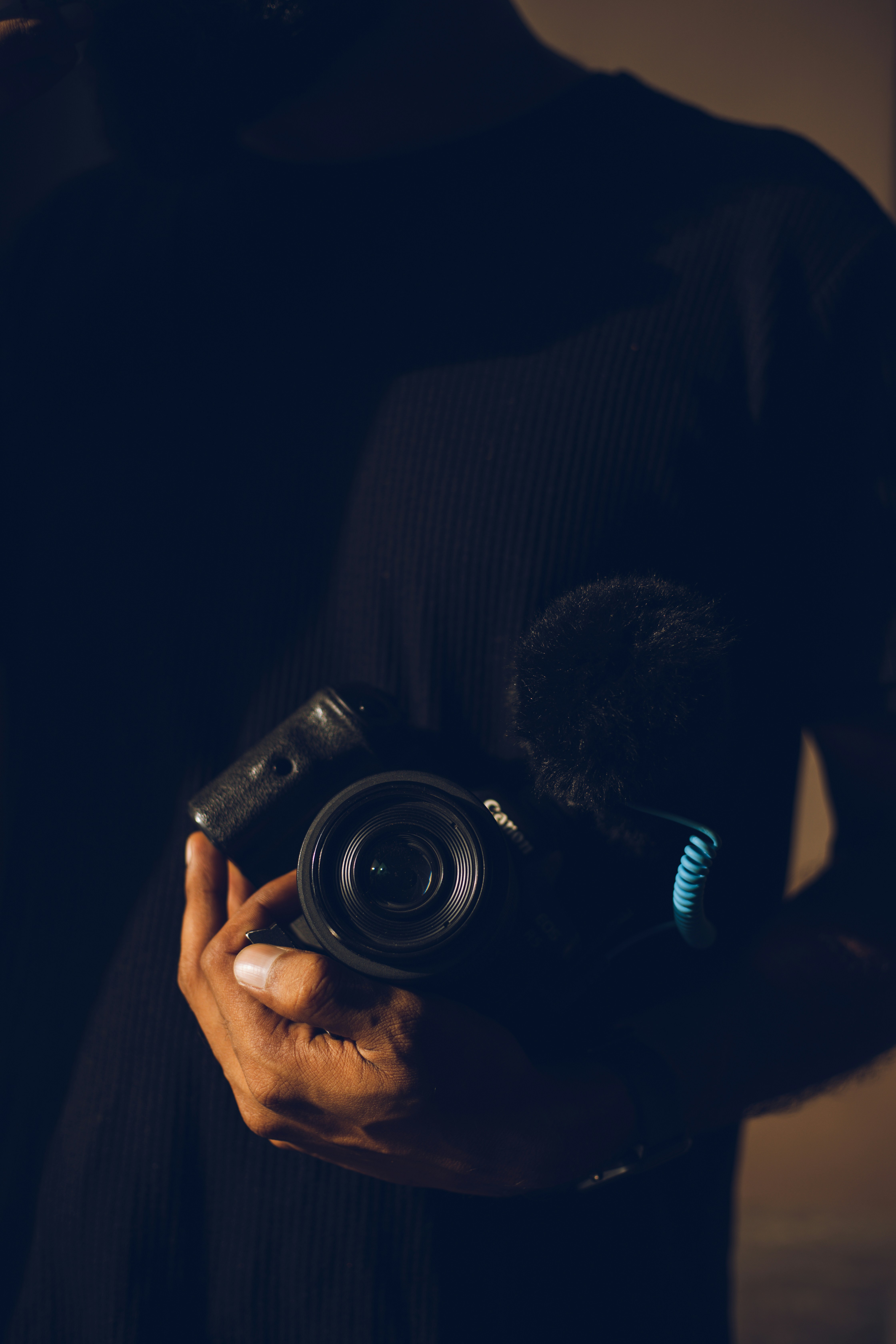 A photographer holds a Canon EOS R camera with a microphone attached, showcasing the tools of visual storytelling. The warm lighting highlights the camera's sleek design.